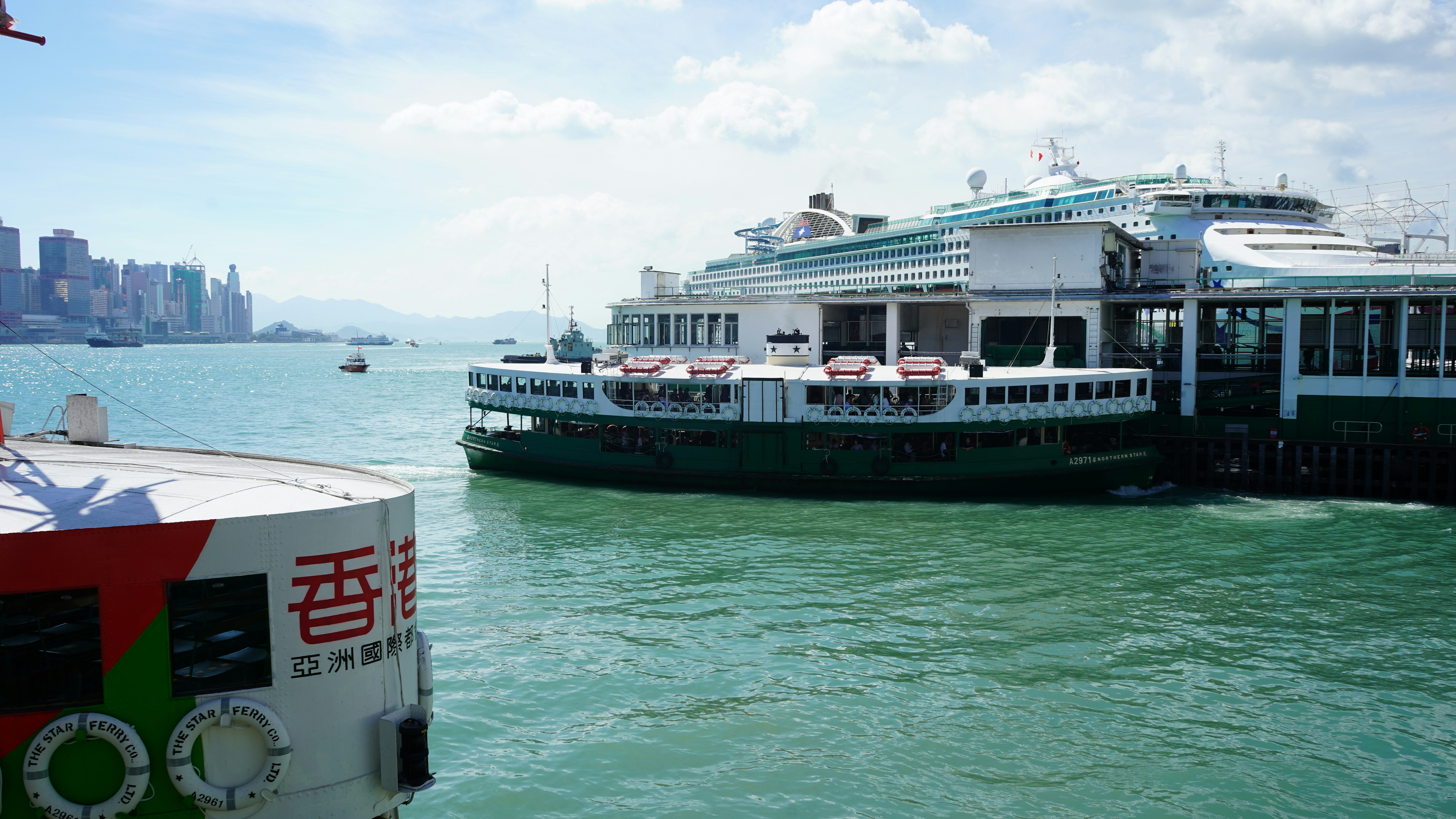 A green-and-white Star Ferry departs from the dock in central Hong Kong, gliding through the glistening waters of Victoria Harbour. The image captures the timeless charm of this iconic ferry system, contrasted against the modern cruise ship terminal and Kowloon skyline in the distance. | Ferry docked in a harbor with city skyline.
