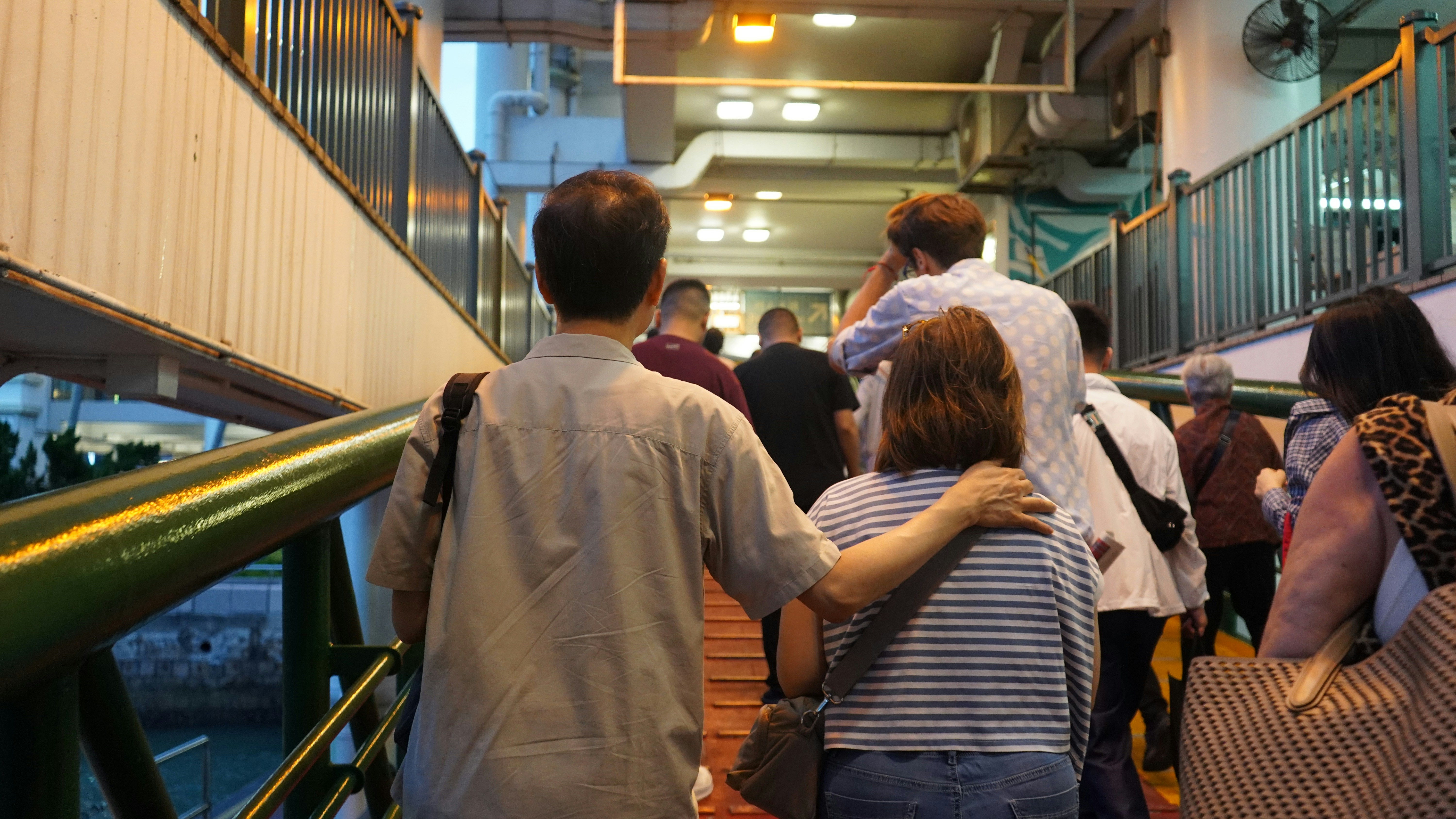 An older couple walks up a harbour ramp on Hong Kong Island, surrounded by commuters. The man’s arm gently rests on the woman’s shoulder in a moment of quiet tenderness. | People walking up a ramp on a ferry