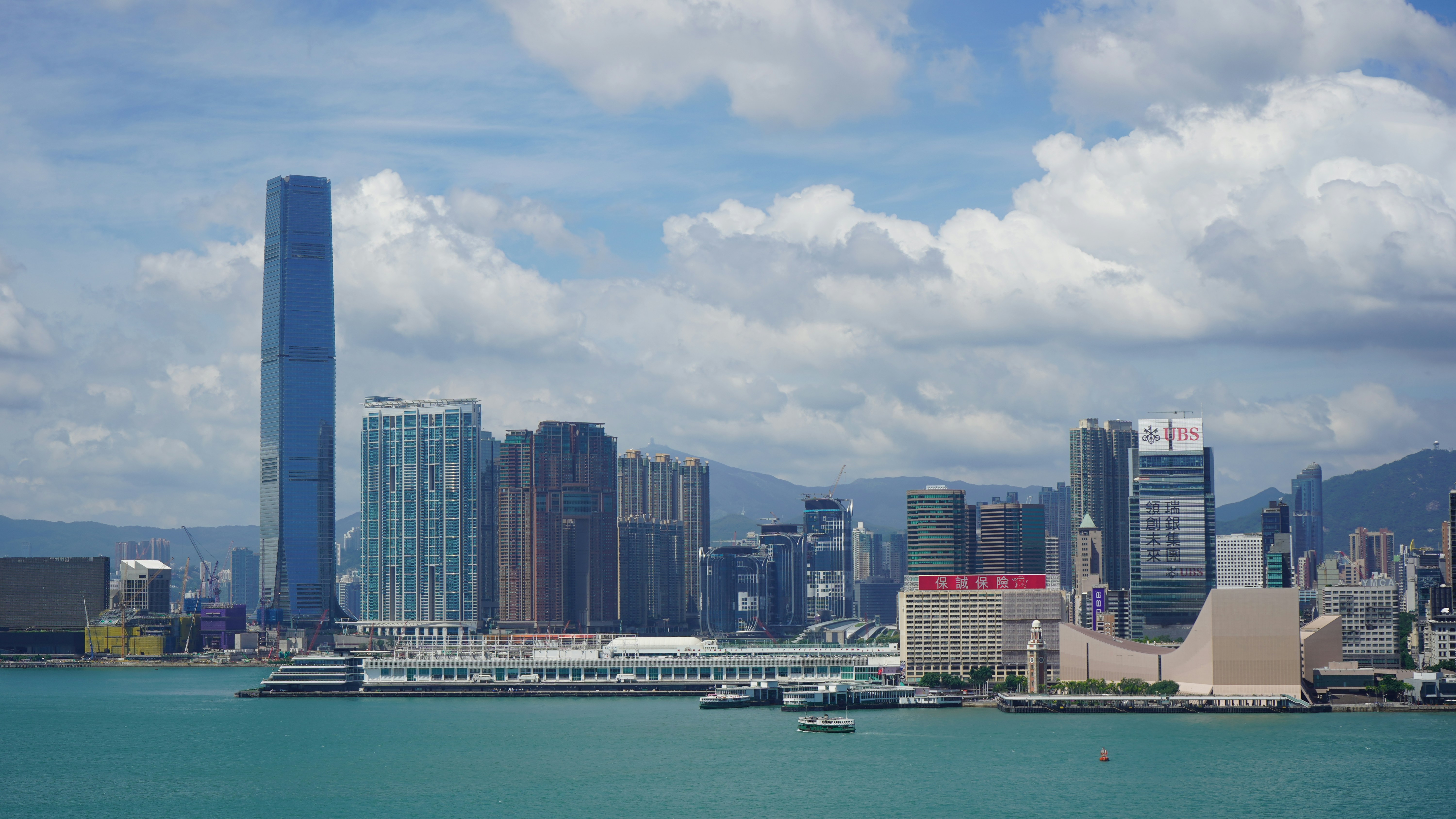 A high-contrast view of Hong Kong’s striking skyline as seen across Victoria Harbour. Towering glass skyscrapers rise above the turquoise sea, with the dramatic International Commerce Centre dominating the scene under bold, cloud-swept skies. | City skyline with tall buildings and a harbor.