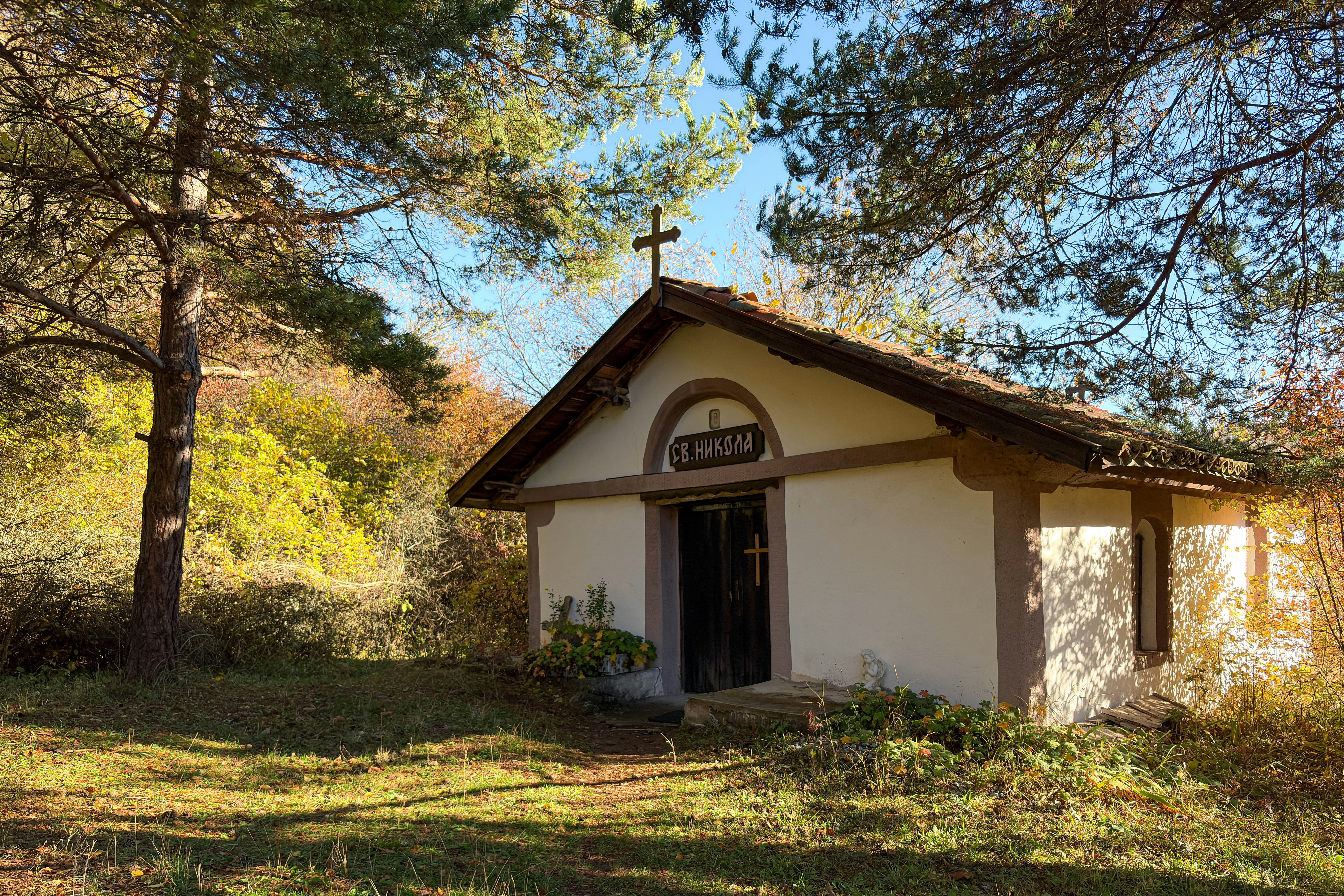 A quaint, whitewashed Orthodox chapel dedicated to St. Nikola stands peacefully in a sunlit forest clearing, framed by pine trees and vibrant golden autumn foliage. /Bailovo, Bulgaria/ | Small chapel with a cross on a hill.