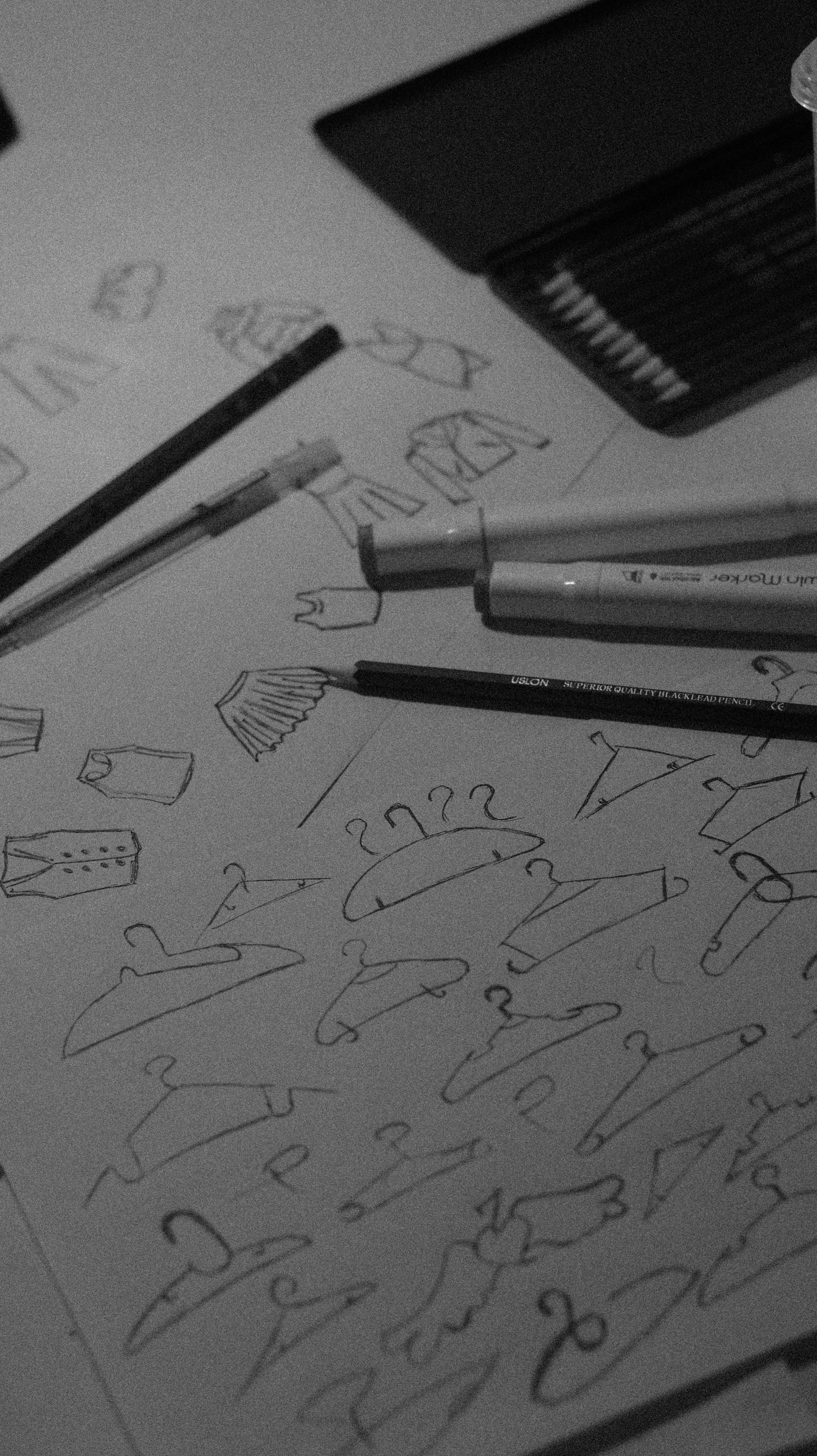 A collection of hand-drawn clothing hangers sketched on paper, surrounded by colored pencils and pens. The monochrome setting emphasizes the artistic process.