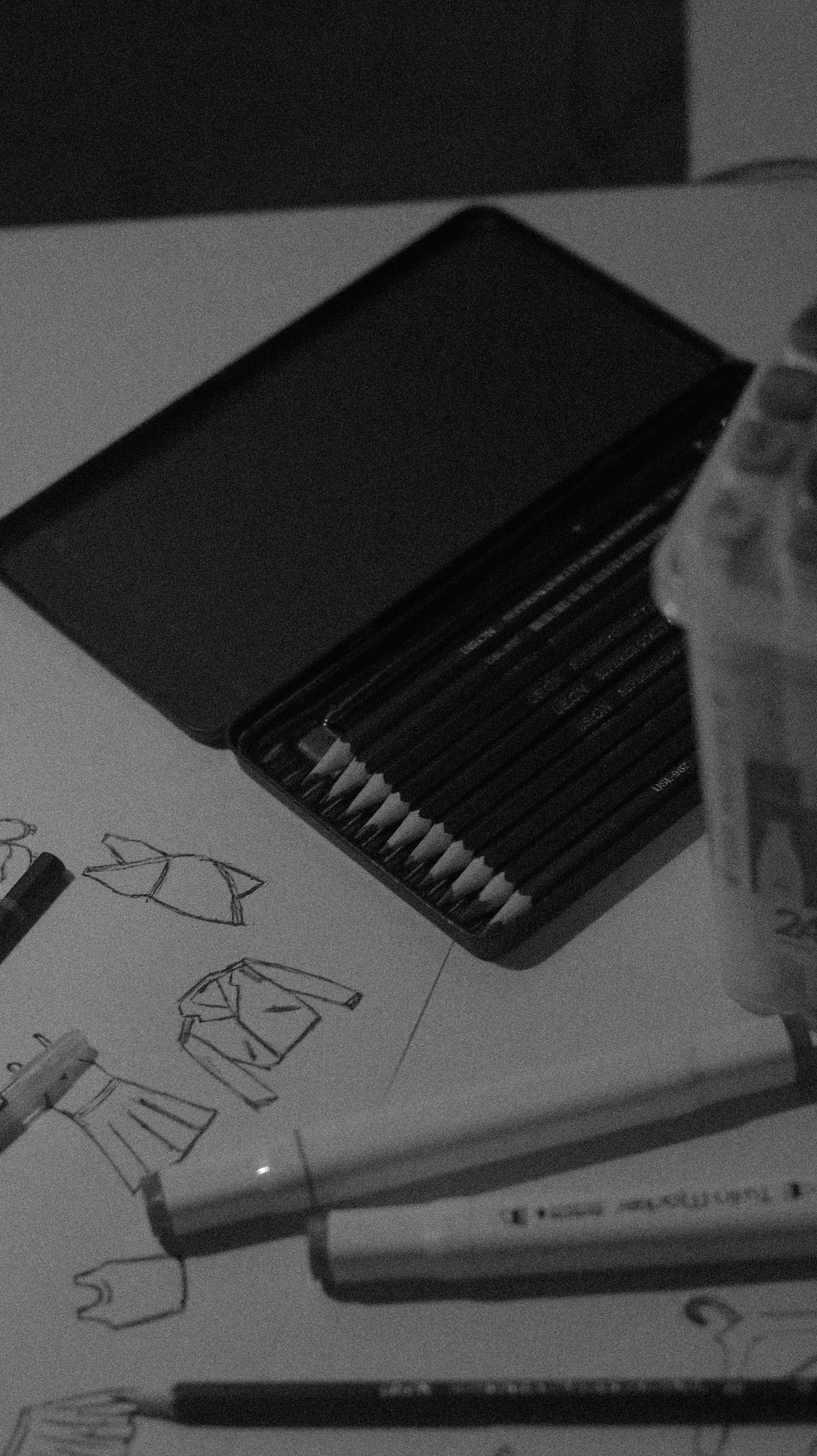 An assortment of colored pencils and sketches of clothing designs arranged on a workspace, highlighting the artistic process. The scene is set in monochrome, emphasizing the creativity involved.