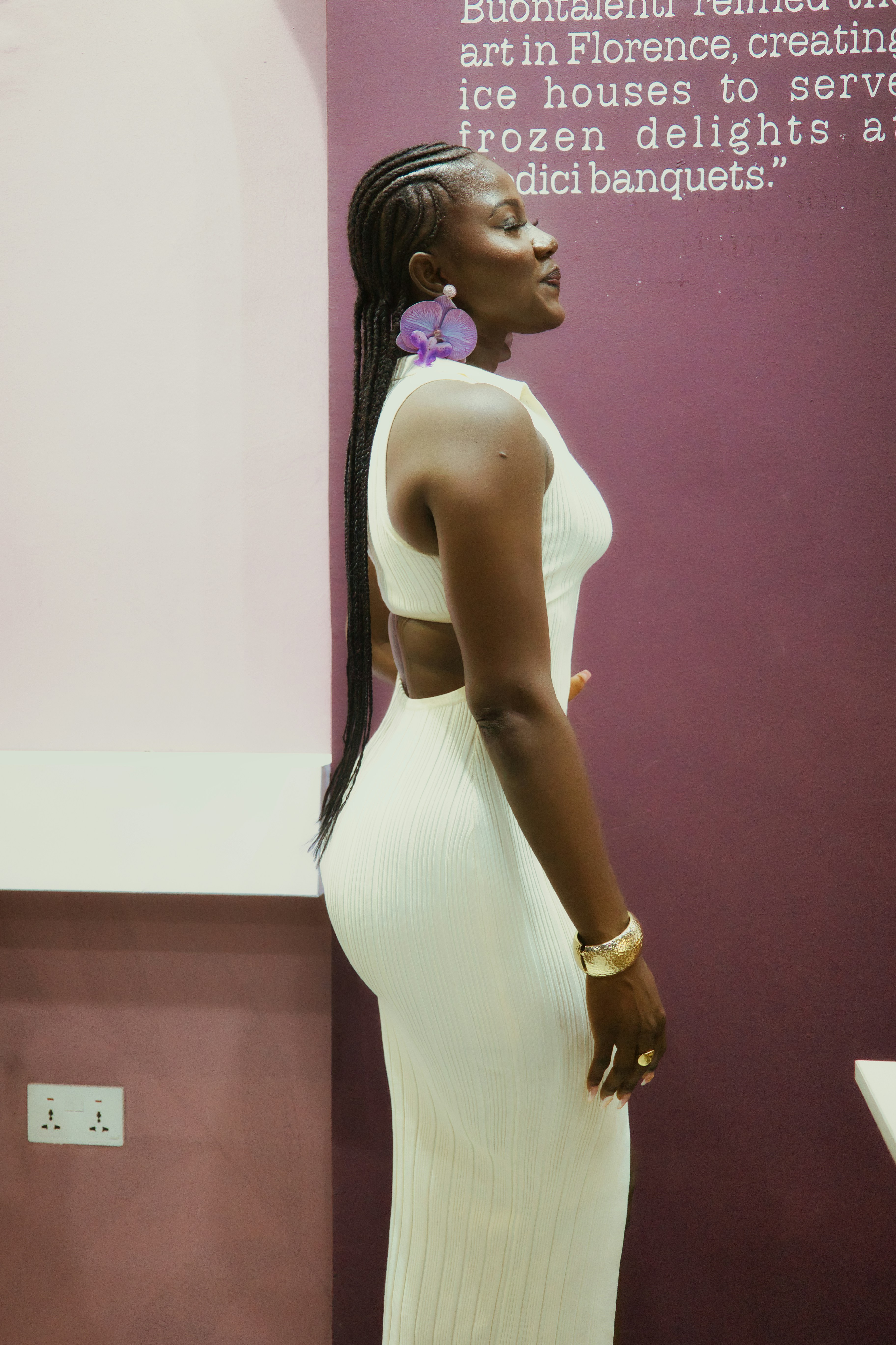 A woman in a fitted white dress poses gracefully against a soft pink wall adorned with text. A purple flower accentuates her hairstyle.