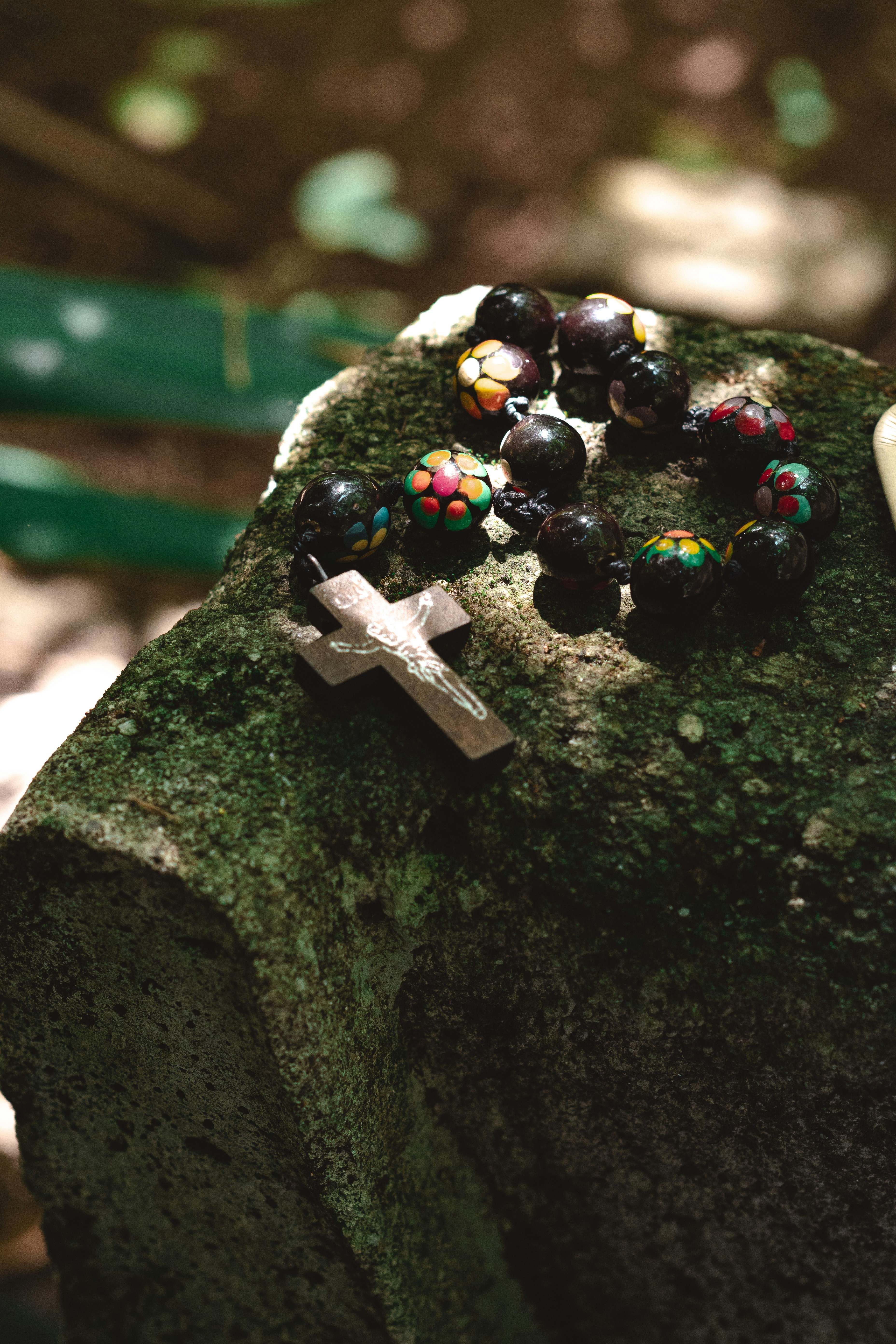 Colorful beaded rosary draped over a moss-covered stone, with a silver cross prominently displayed. Natural light highlights the textures and colors.