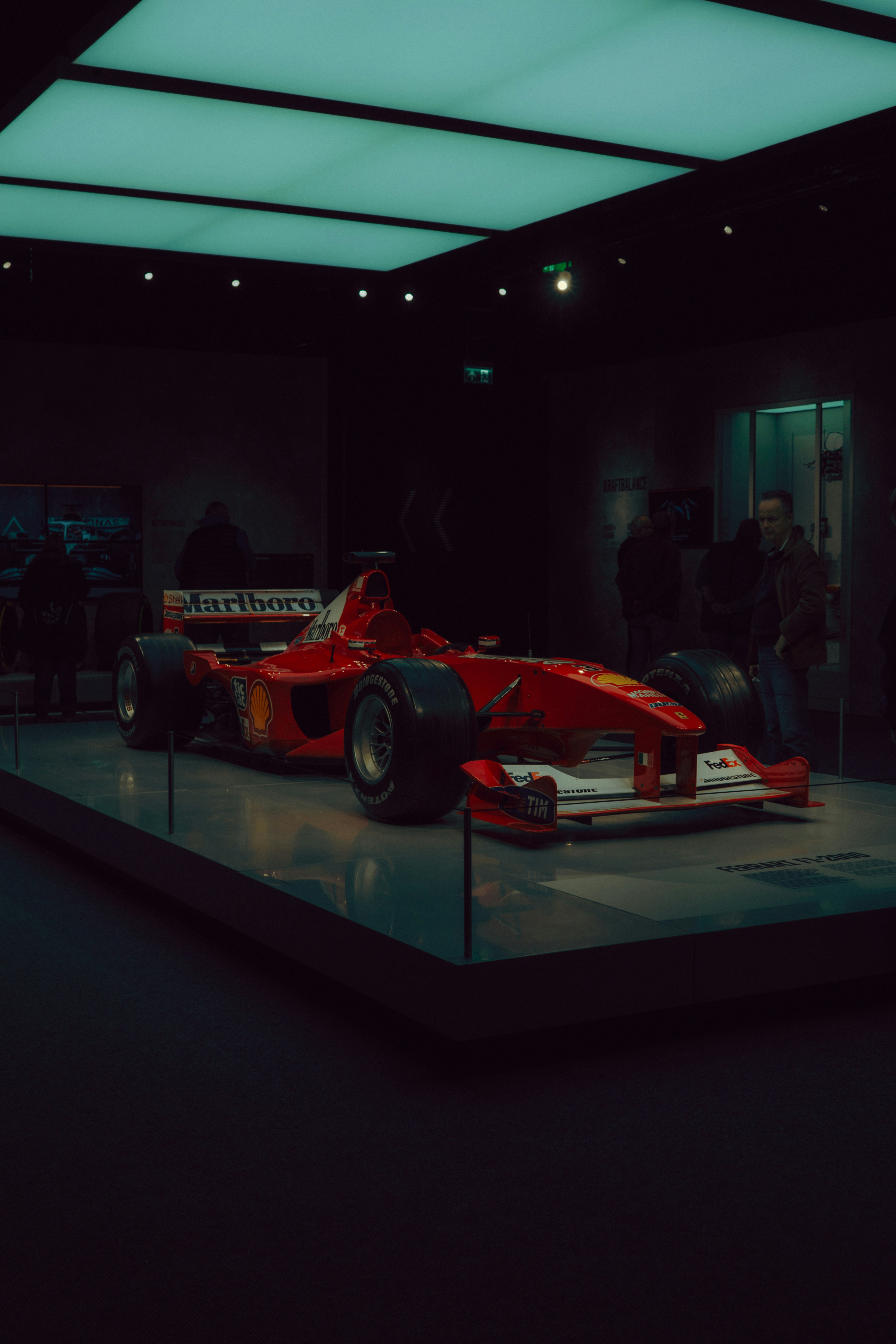A classic red race car showcased in a modern exhibit, surrounded by intrigued visitors. The sleek design highlights its racing heritage.