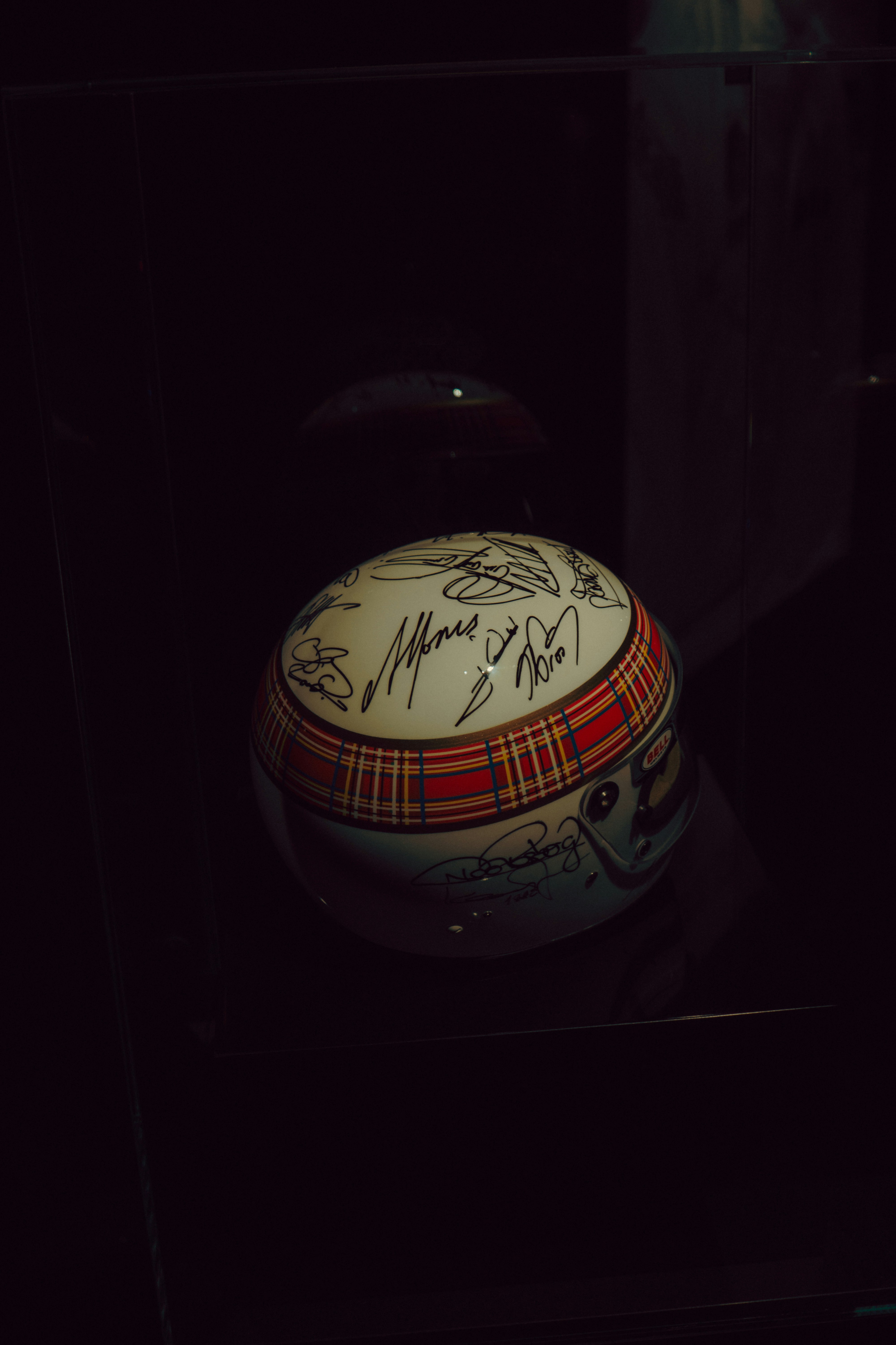 A vintage racing helmet adorned with various signatures, displayed in a glass case. The helmet's design features a vibrant plaid pattern.