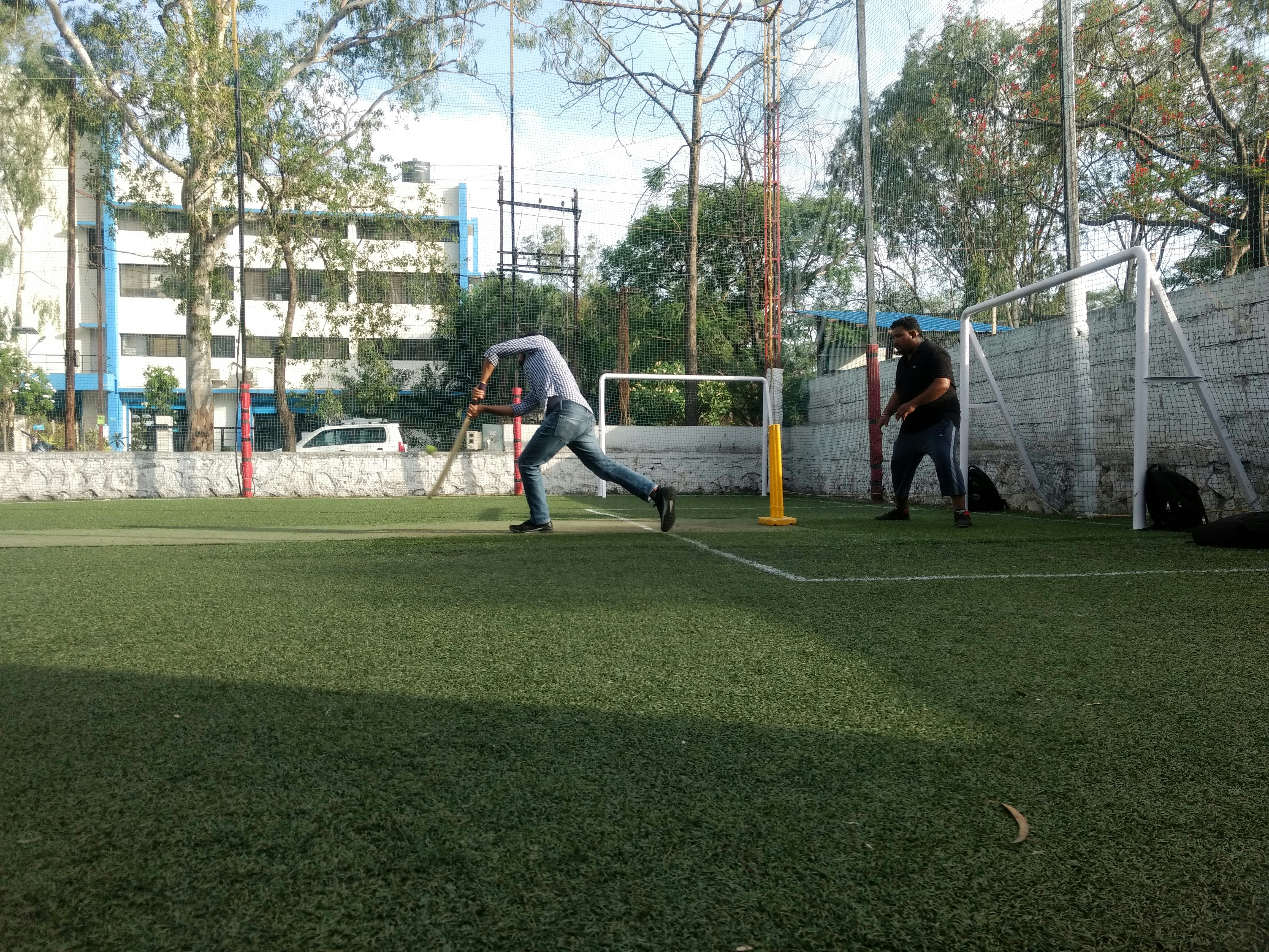 Two players engaged in cricket practice on a synthetic turf field surrounded by trees and urban structures.