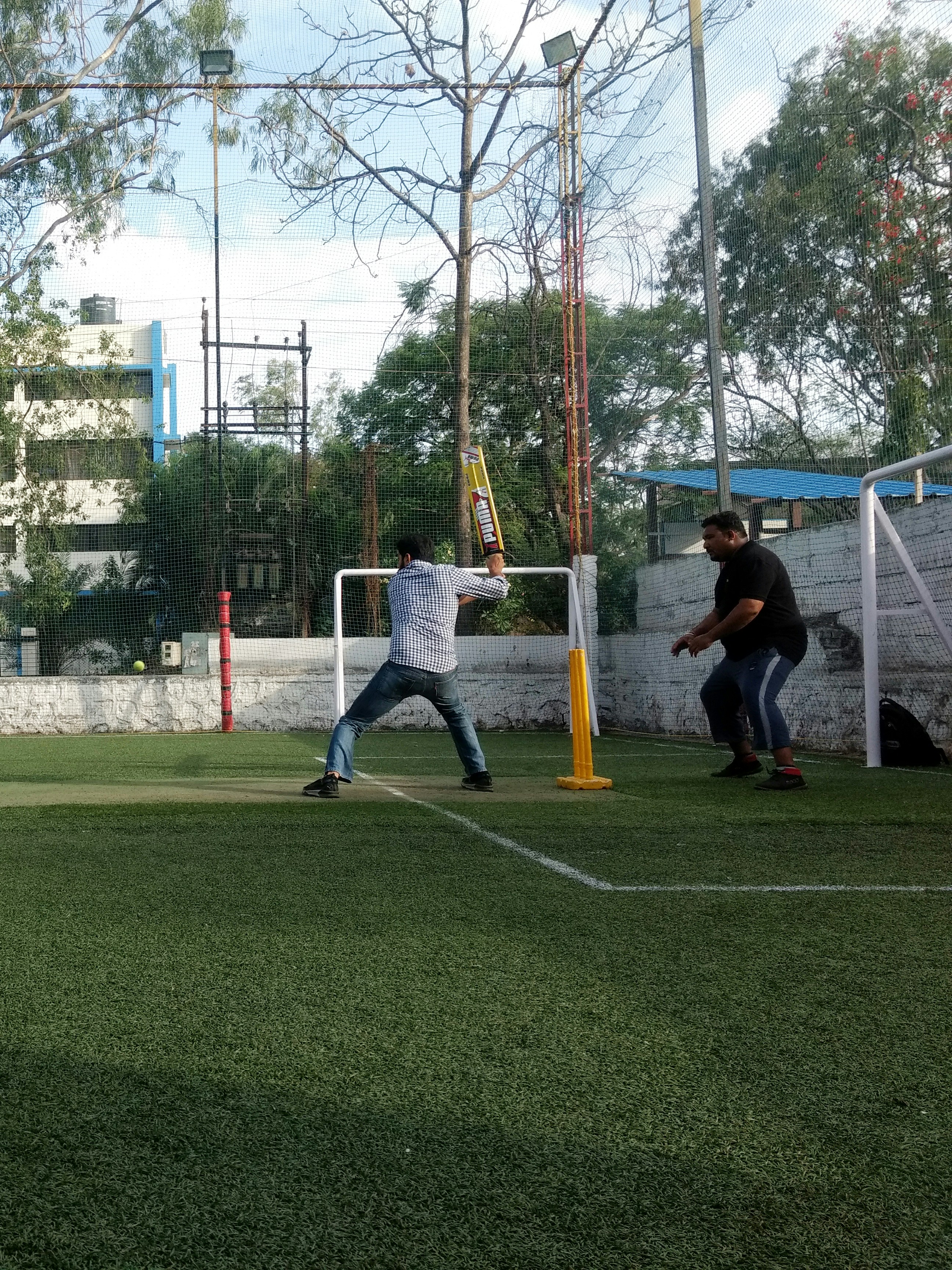 Two players engaged in a spirited game of cricket on a well-maintained turf field, with a backdrop of greenery and urban structures.
