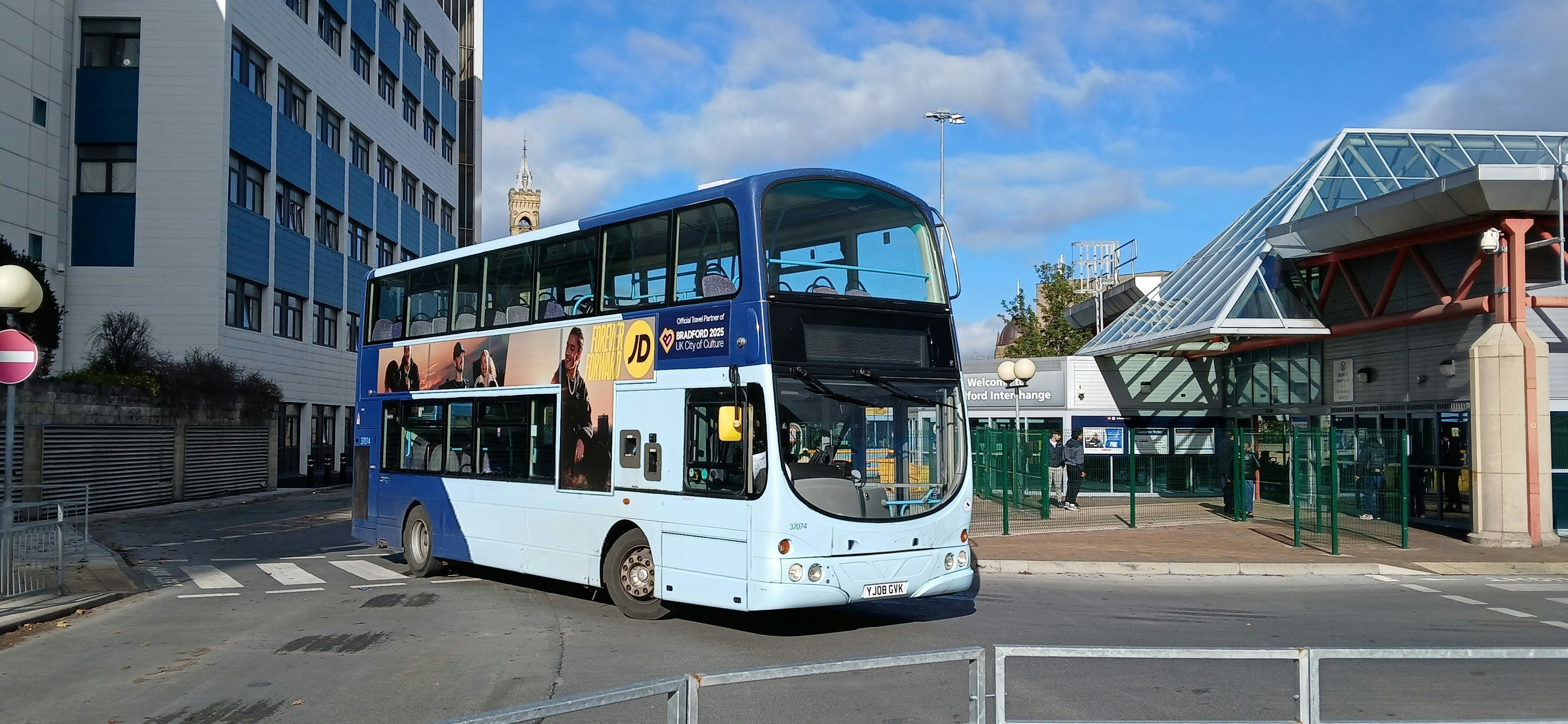 A two-tone blue liveried Bradford bus in Bradford Interchange, West Yorkshire, England, United Kingdom. Taken on Friday 24th October 2025. | A double-decker bus turns a corner near a modern building.