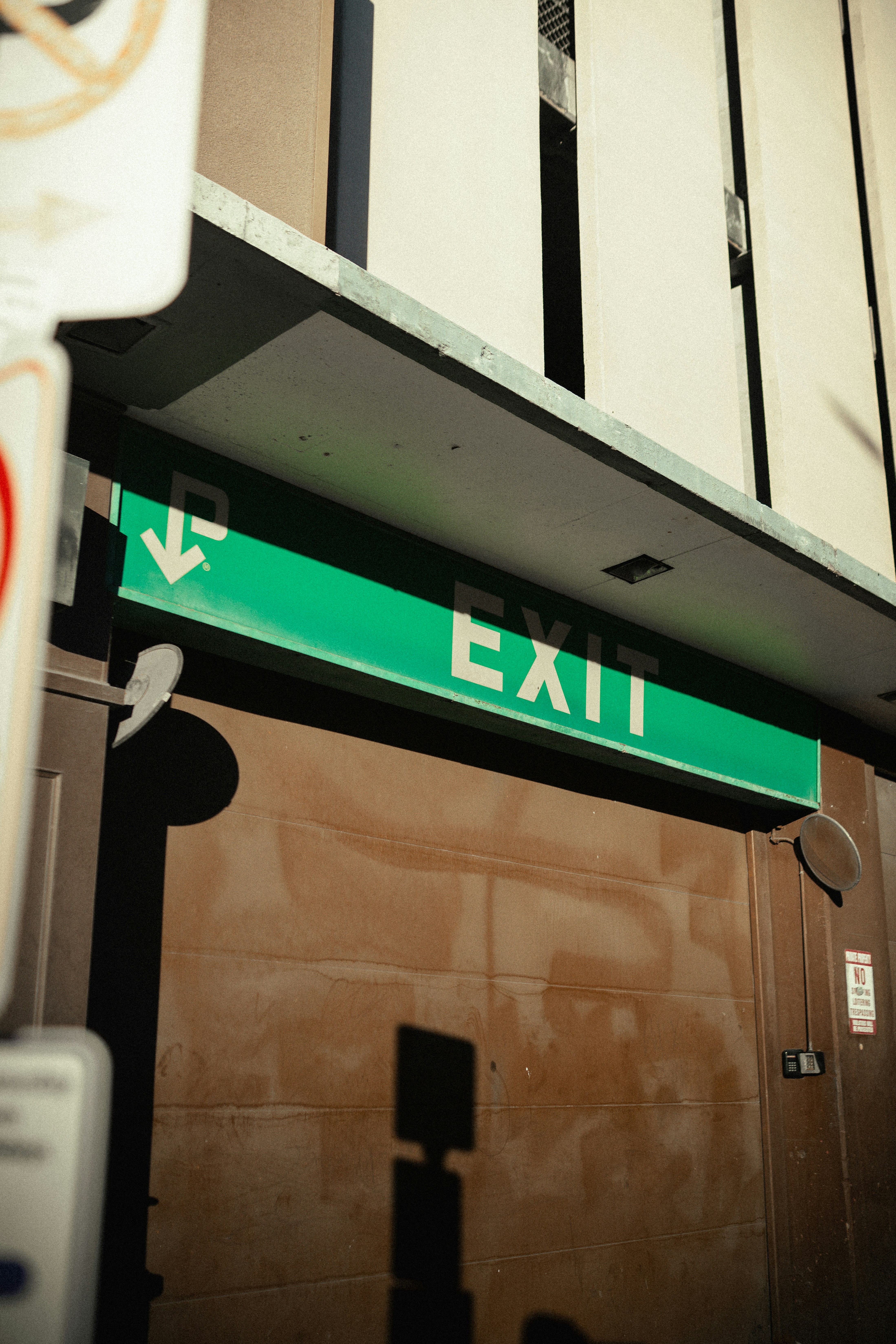 Bright green exit sign illuminated against a textured brown wall, guiding pedestrians through an urban environment.