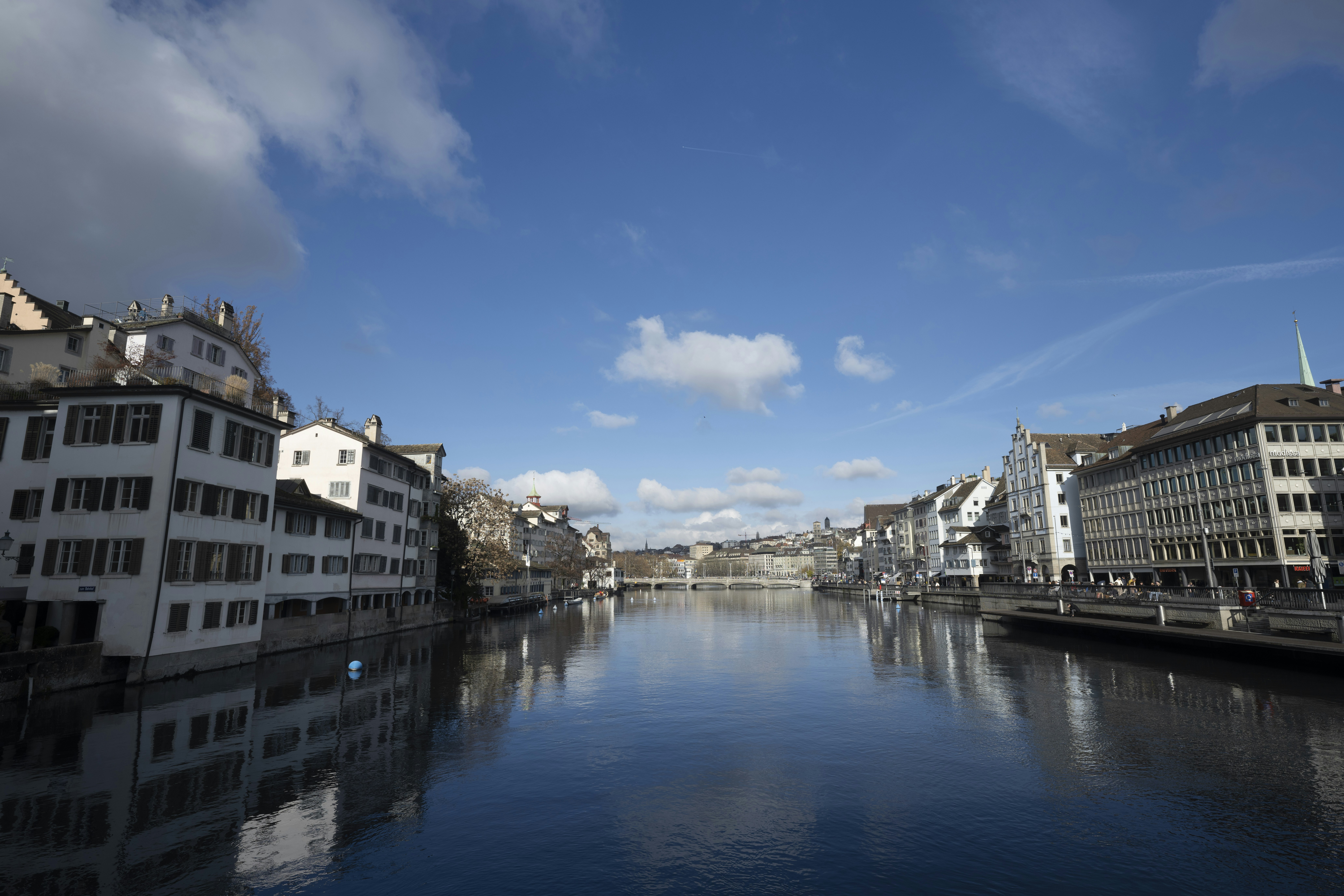 Serene view of Zurich's waterfront, showcasing historic buildings lining the riverbank under a partly cloudy sky.