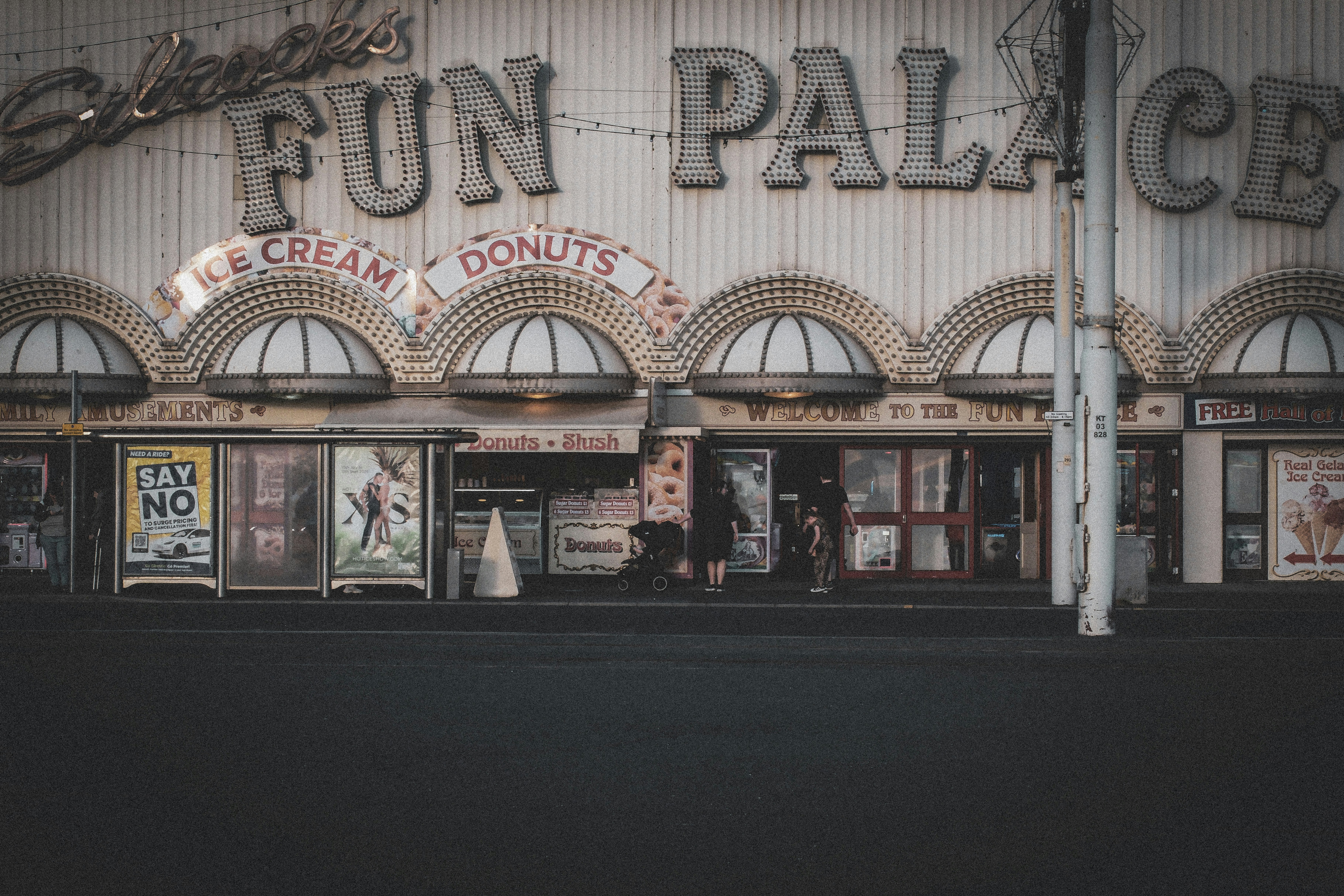 Moody urban night scene of a retro funfair facade with neon lights. The image has a cinematic, arthouse atmosphere, reminiscent of a film still. | Fun palace amusement arcade with ice cream and donuts
