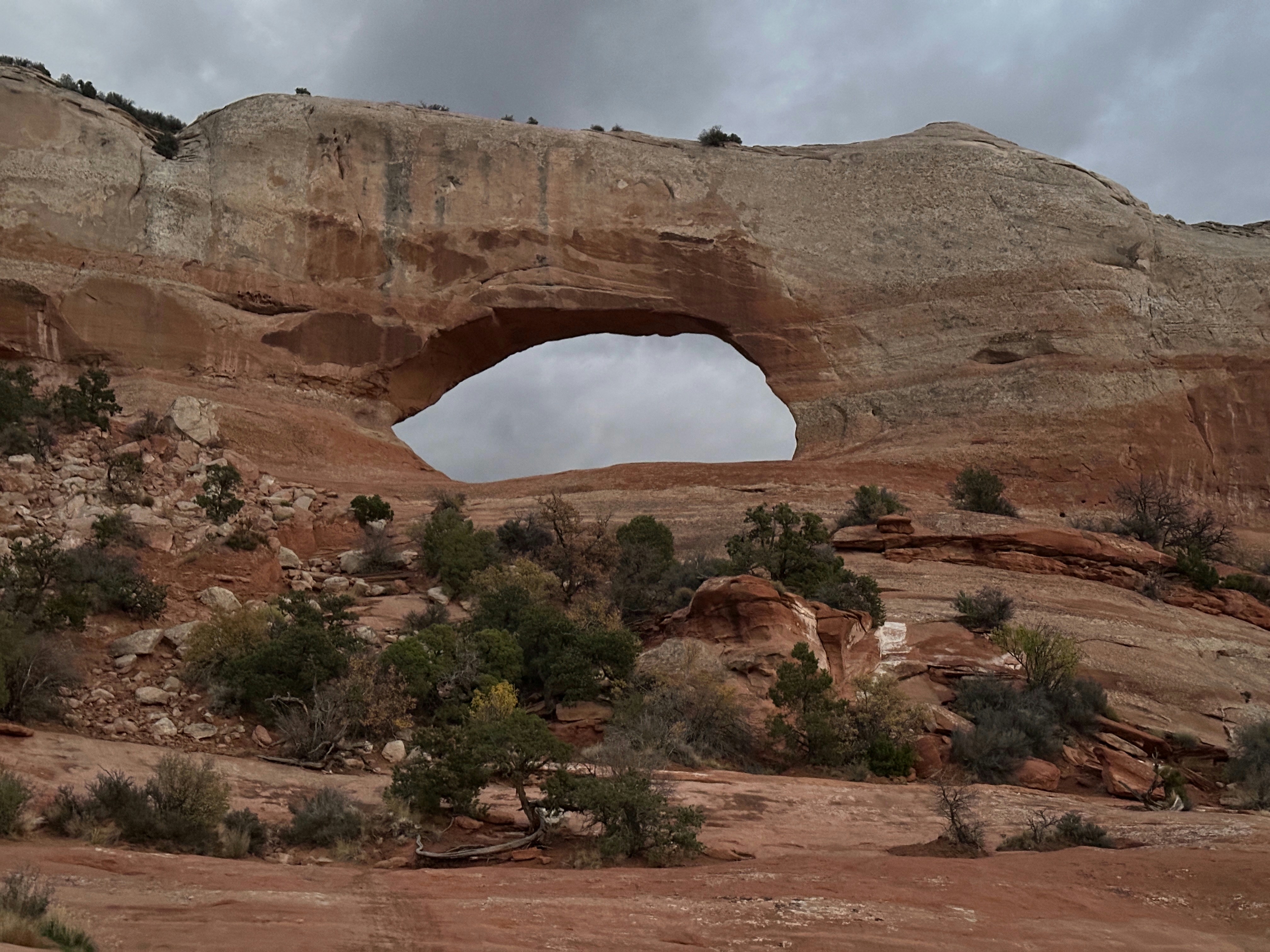 Natural rock arch framed by rugged terrain under a cloudy sky. Lush vegetation contrasts with the earthy tones of the landscape.