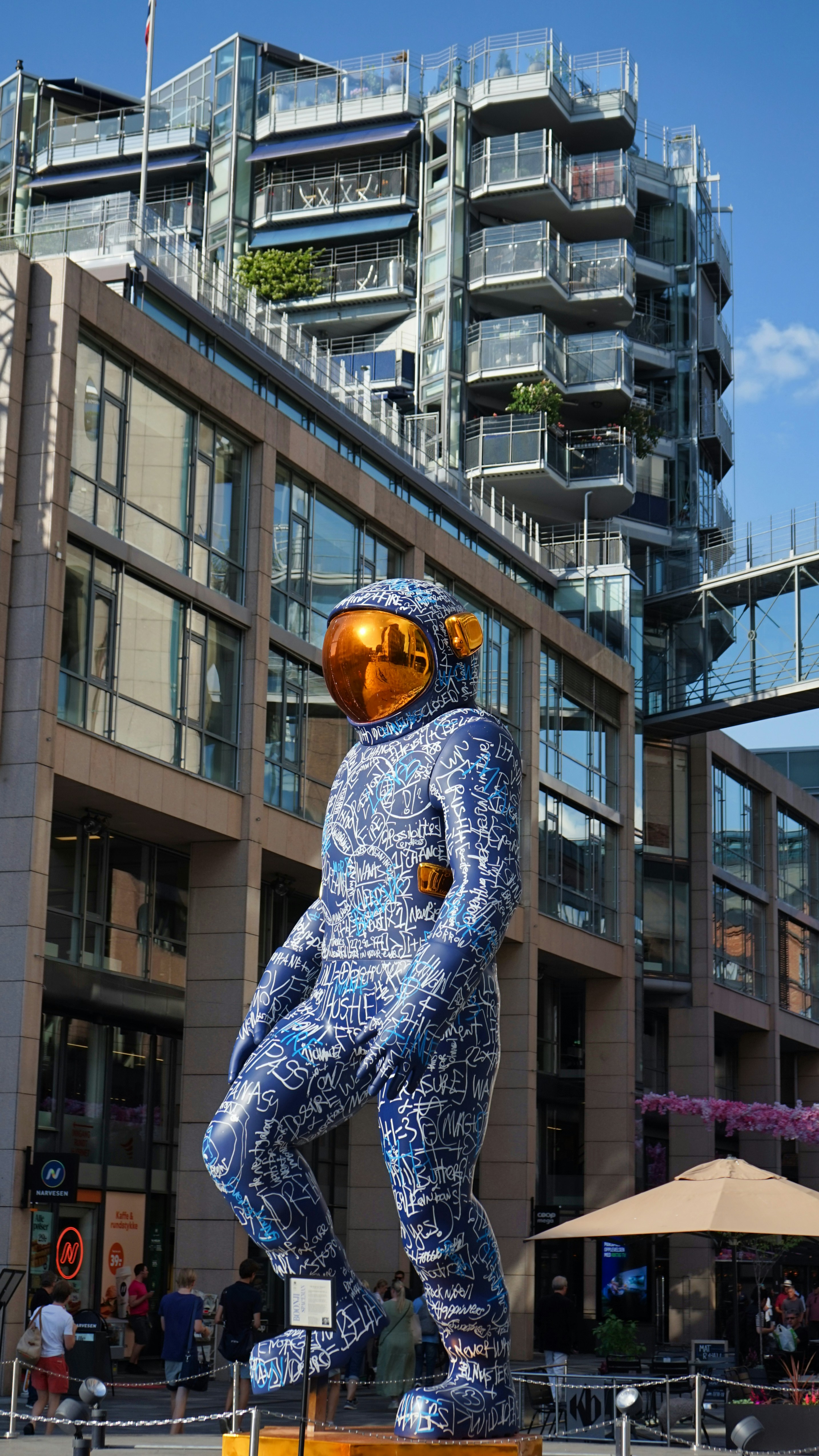 Vibrant astronaut sculpture adorned with intricate blue graffiti stands prominently in an urban plaza, surrounded by modern architecture and curious onlookers.
