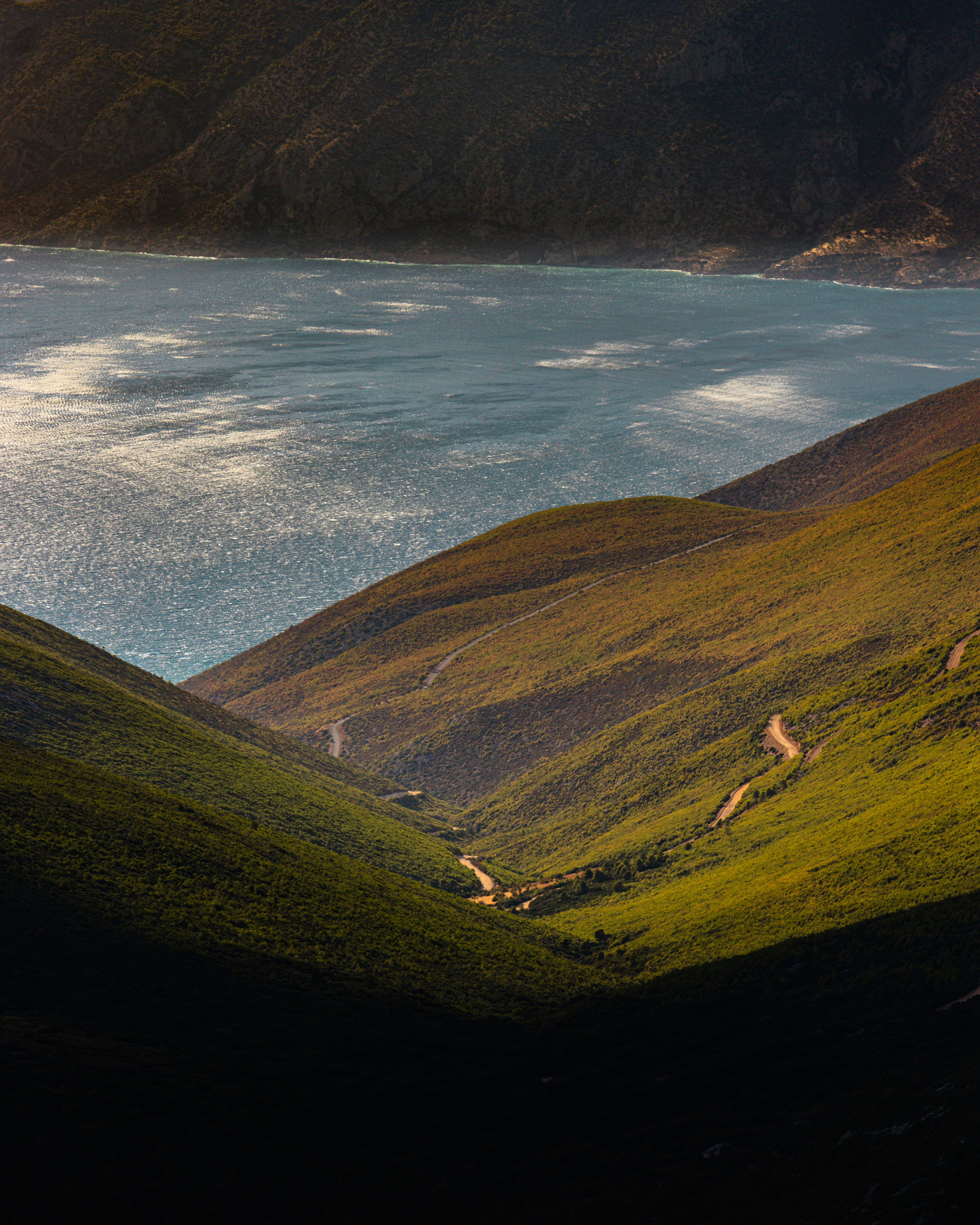 Sunlit green hills with a winding road and blue water.