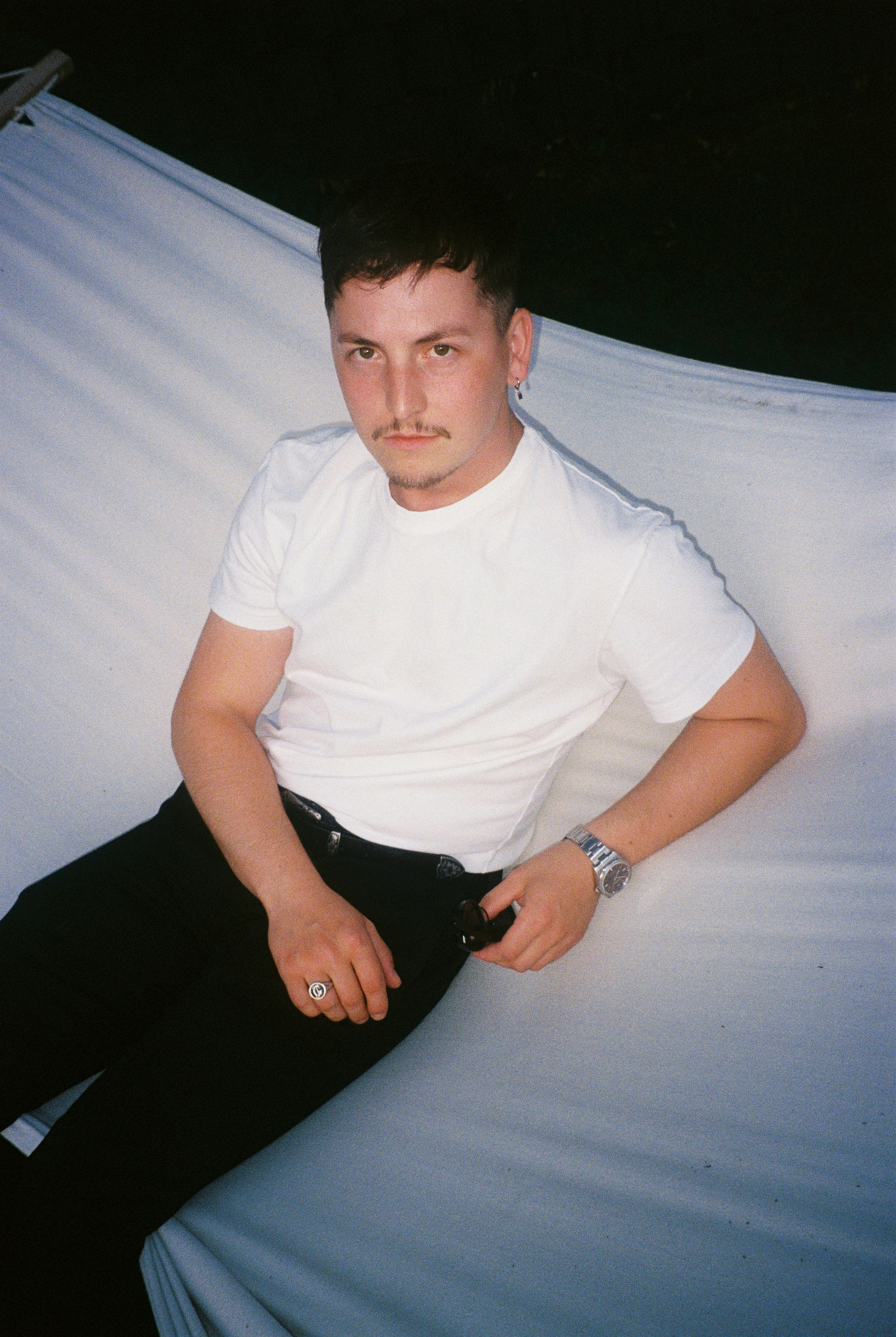 A young man lounging comfortably on a hammock, wearing a white t-shirt and black pants, with a watch and ring visible. The setting conveys a casual, laid-back atmosphere.