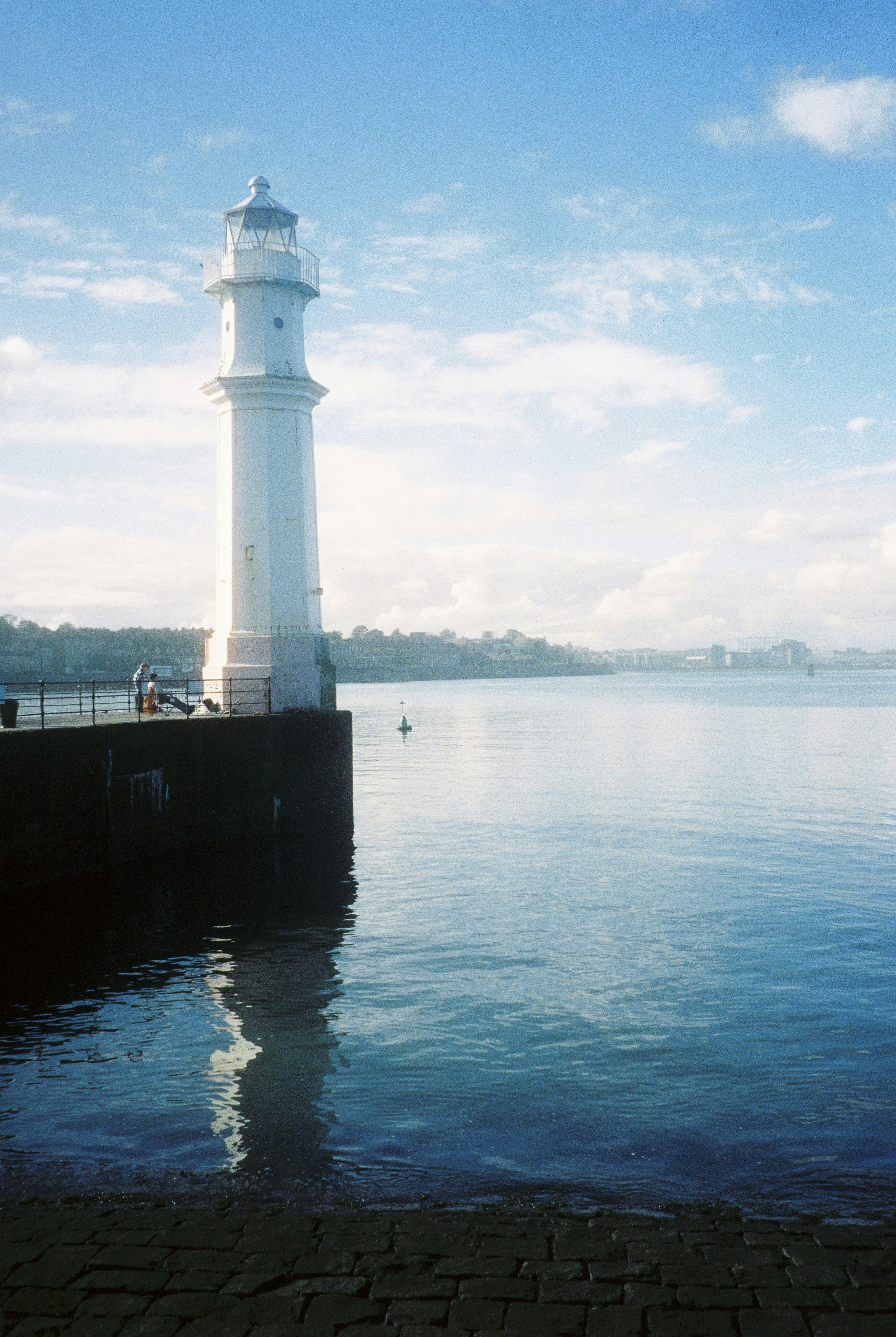 A tall lighthouse stands proudly on the edge of a serene harbor, reflecting in the calm waters beneath a clear blue sky.