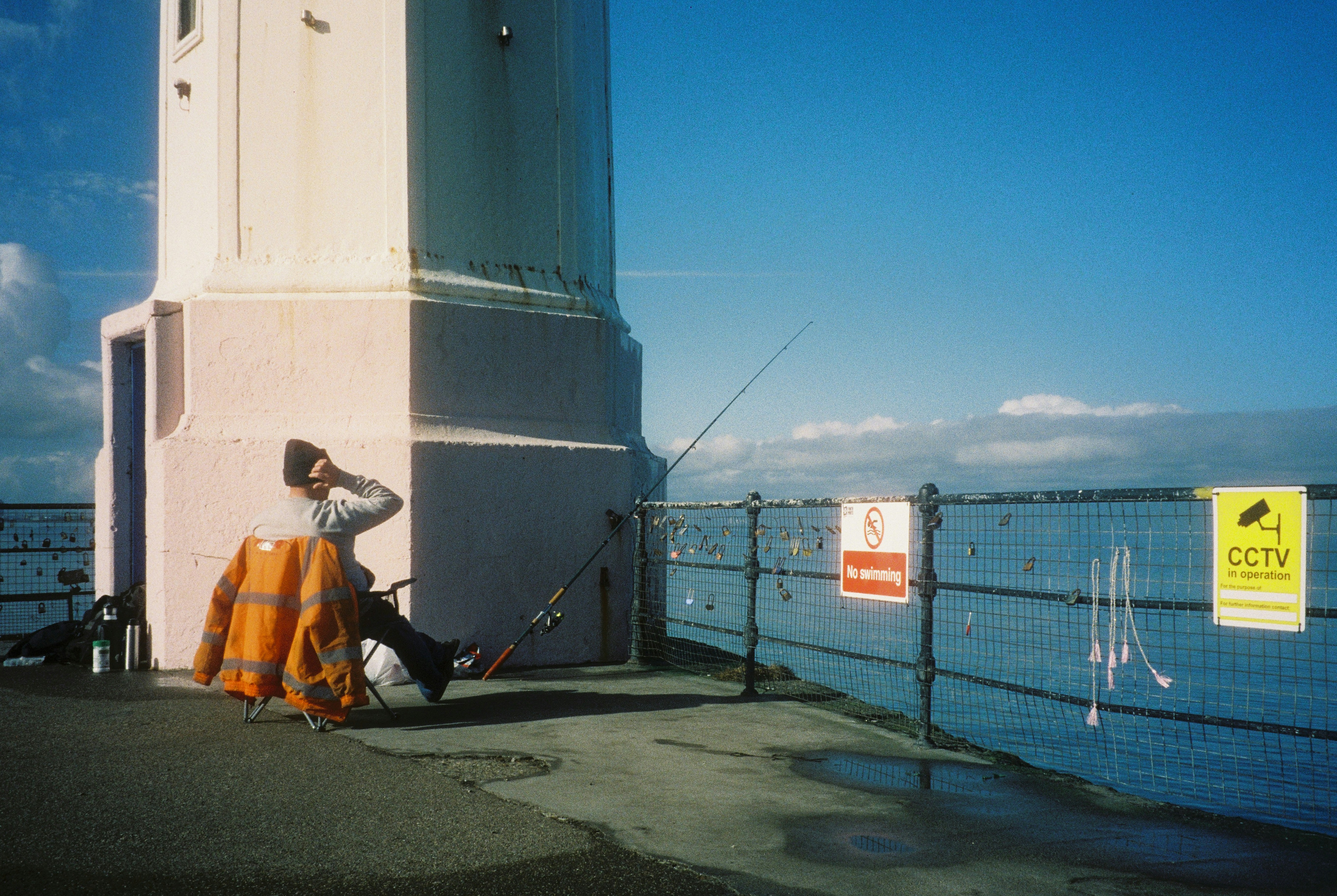 Fisherman relaxing in a chair beside a lighthouse, casting a line into the tranquil waters, with safety signs visible nearby.