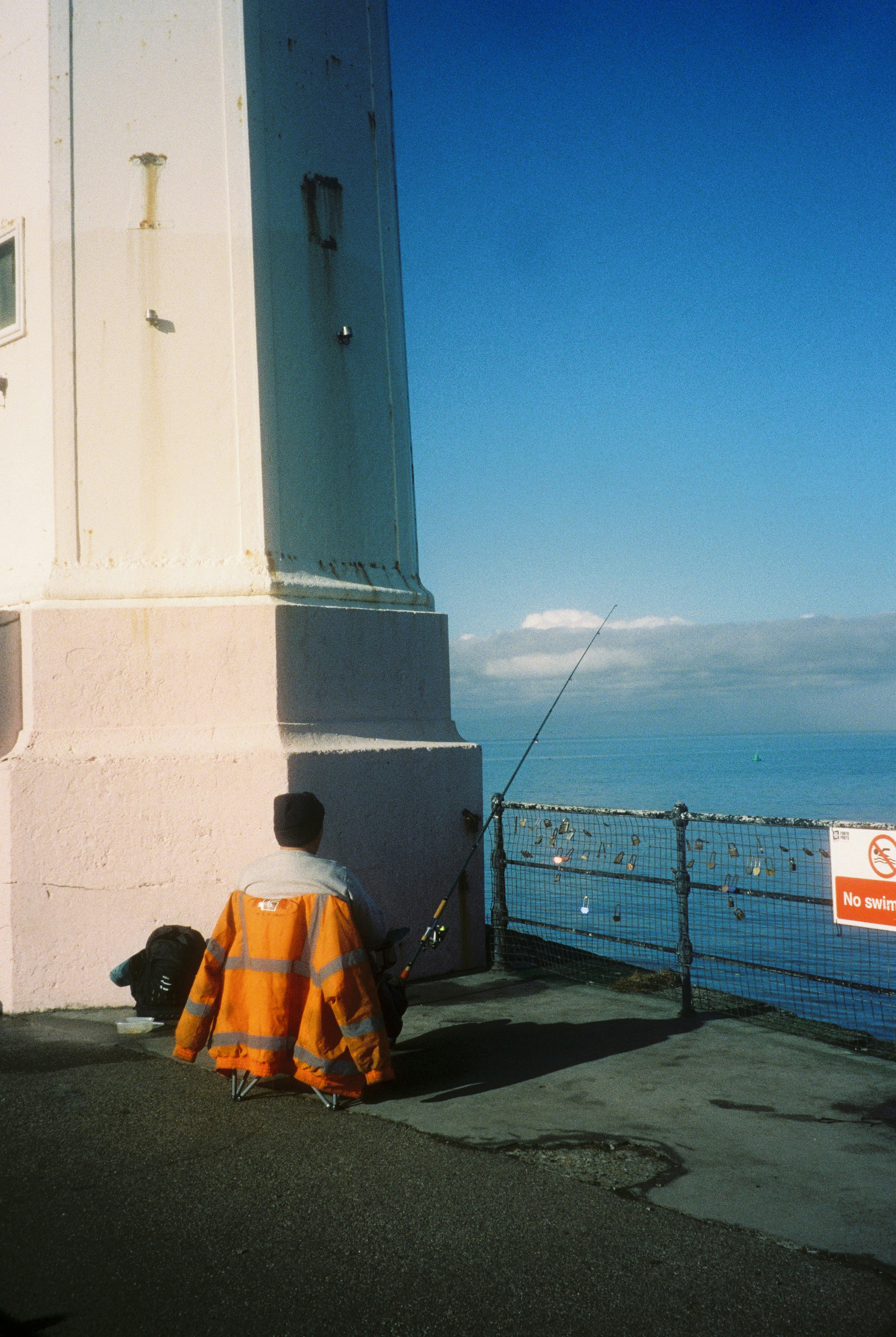 Fisherman in an orange jacket sits calmly by the water, casting a line near a lighthouse. A serene coastal scene unfolds behind him.