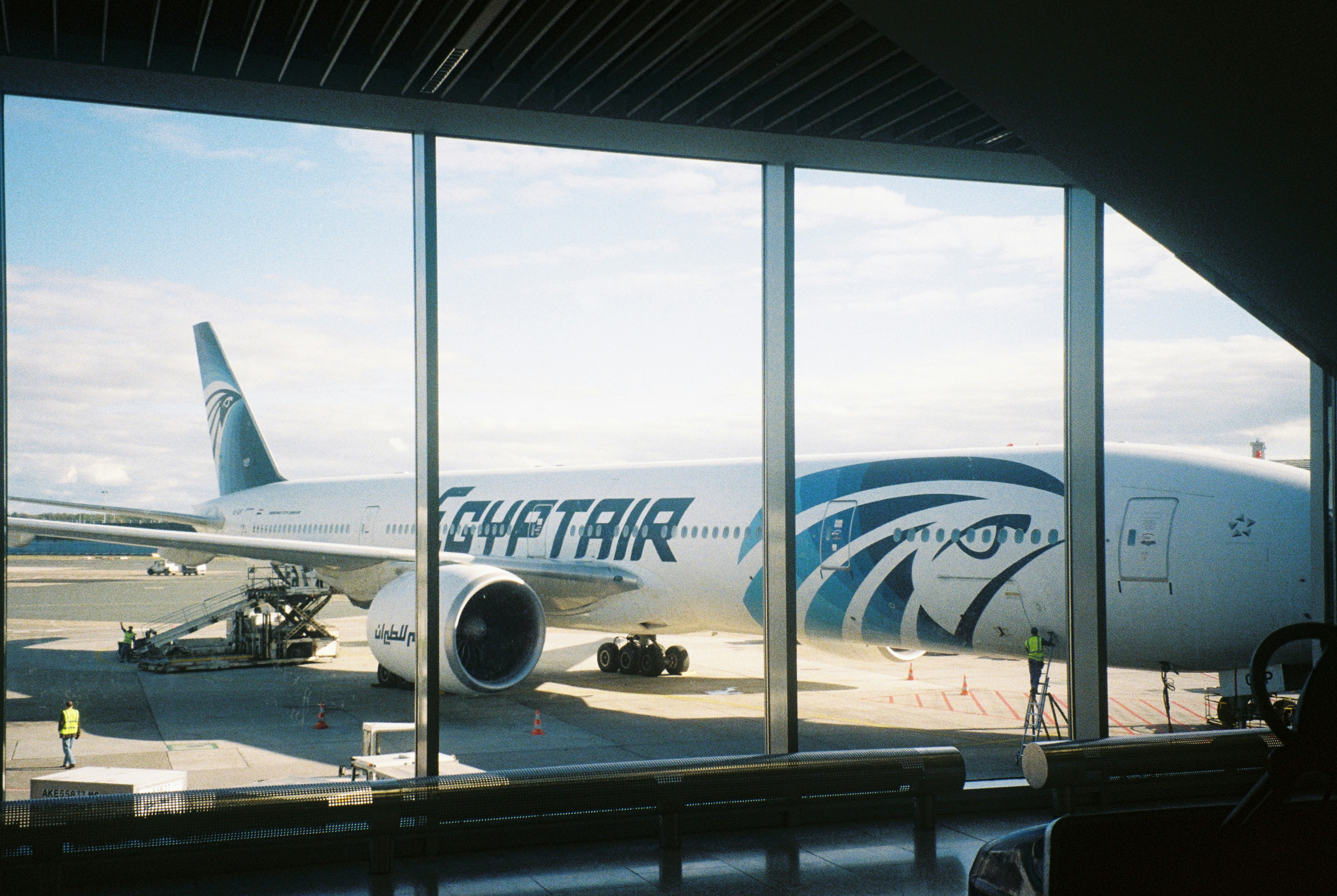 EgyptAir Boeing 777 parked at the airport, showcasing its distinctive livery through large glass windows. Ground crew prepares for the next flight.