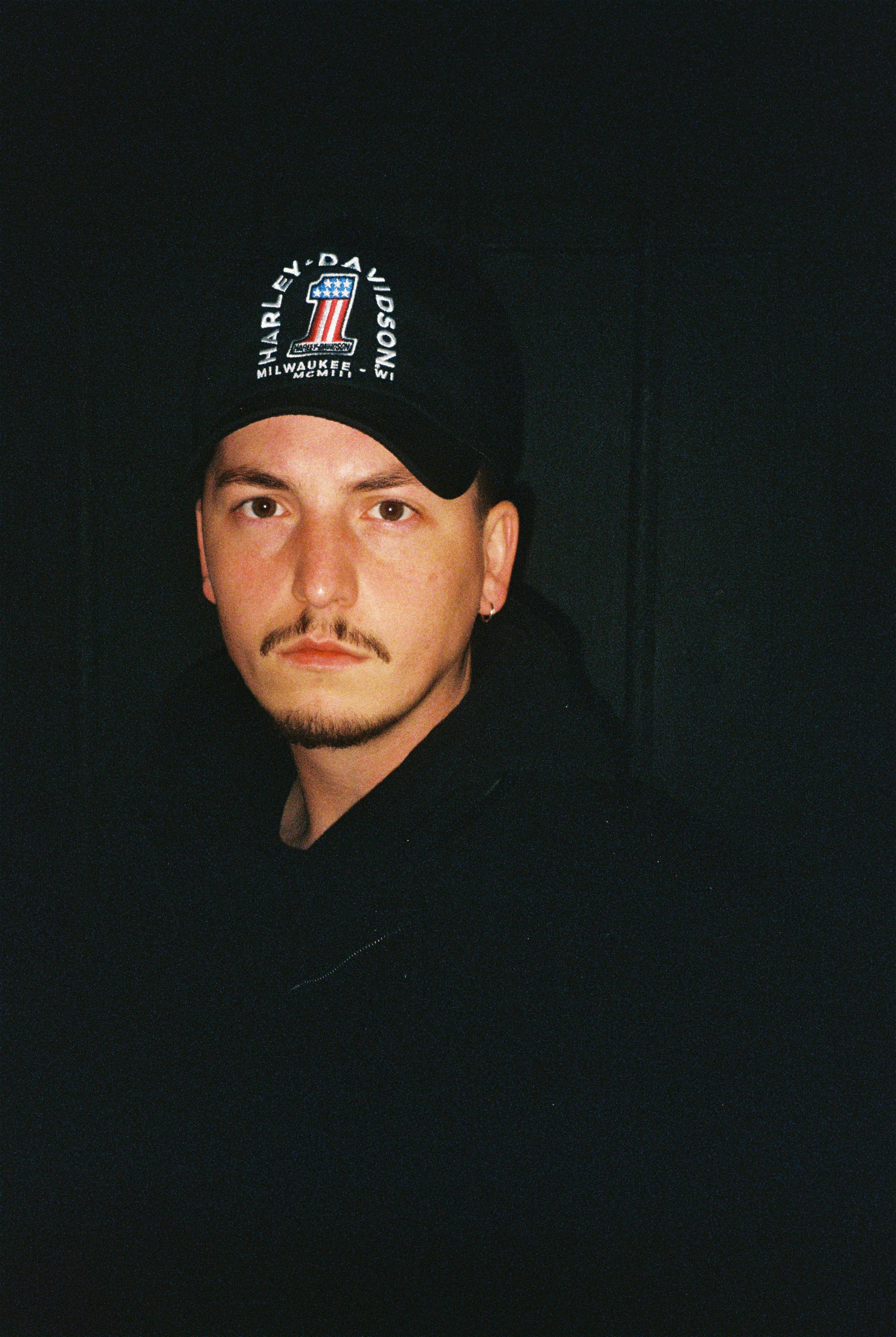 Man with a focused expression wearing a black hoodie and a Harley-Davidson cap against a dark background.