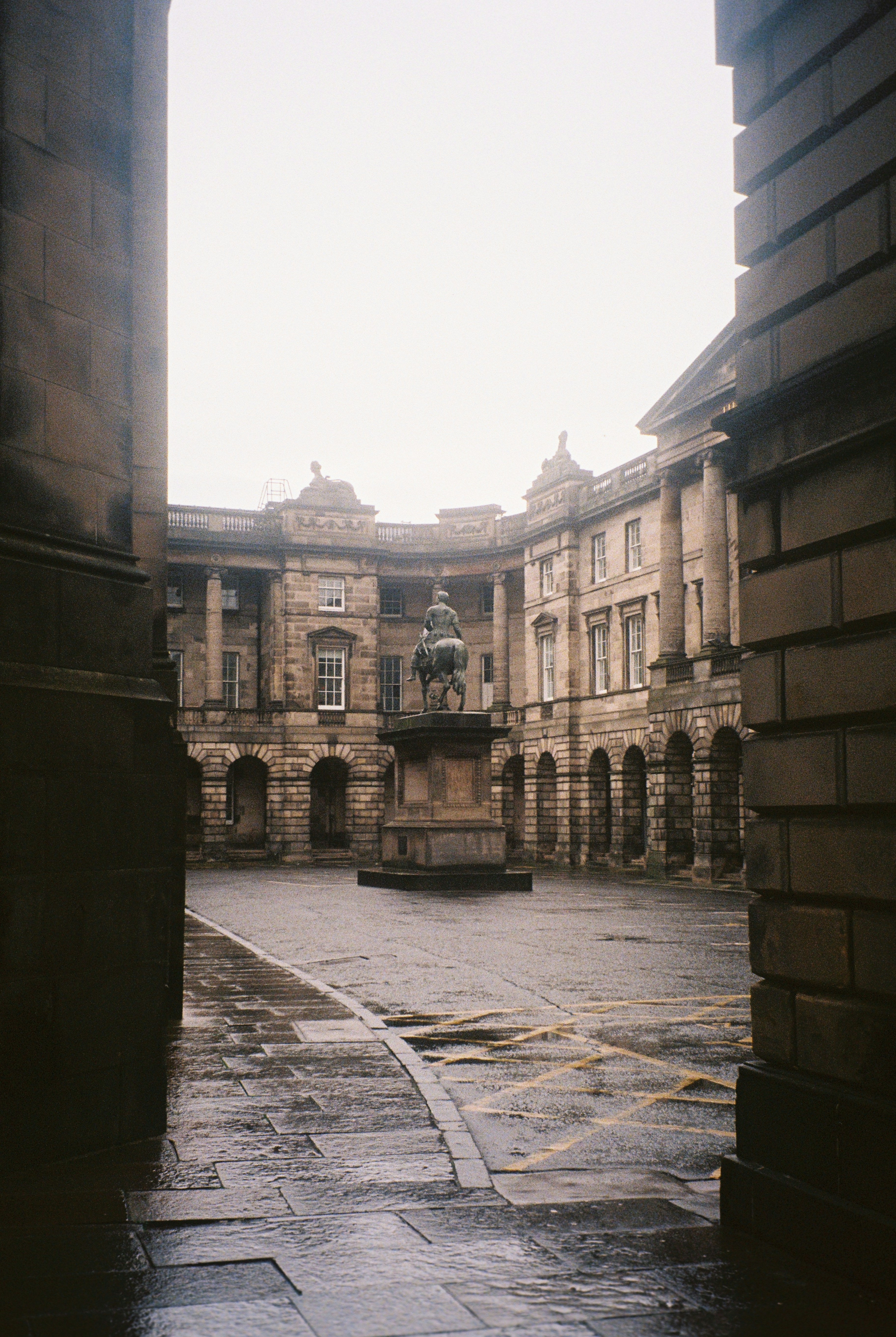 A statue of a mounted figure stands in a courtyard surrounded by classical architecture, captured on a rainy day.