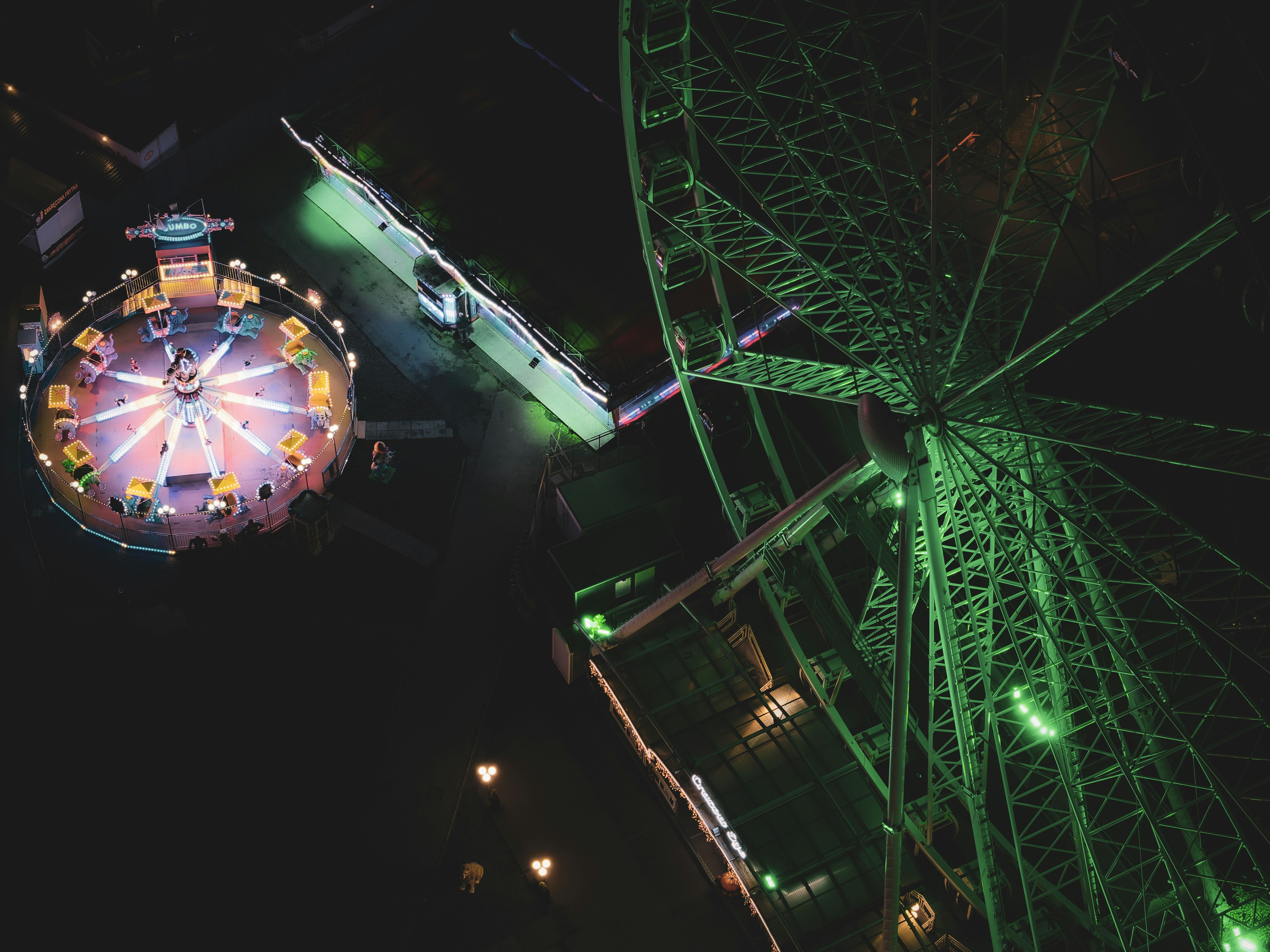 Aerial view of a vibrant amusement park scene featuring a spinning ride surrounded by colorful lights, with a towering Ferris wheel illuminated in green.