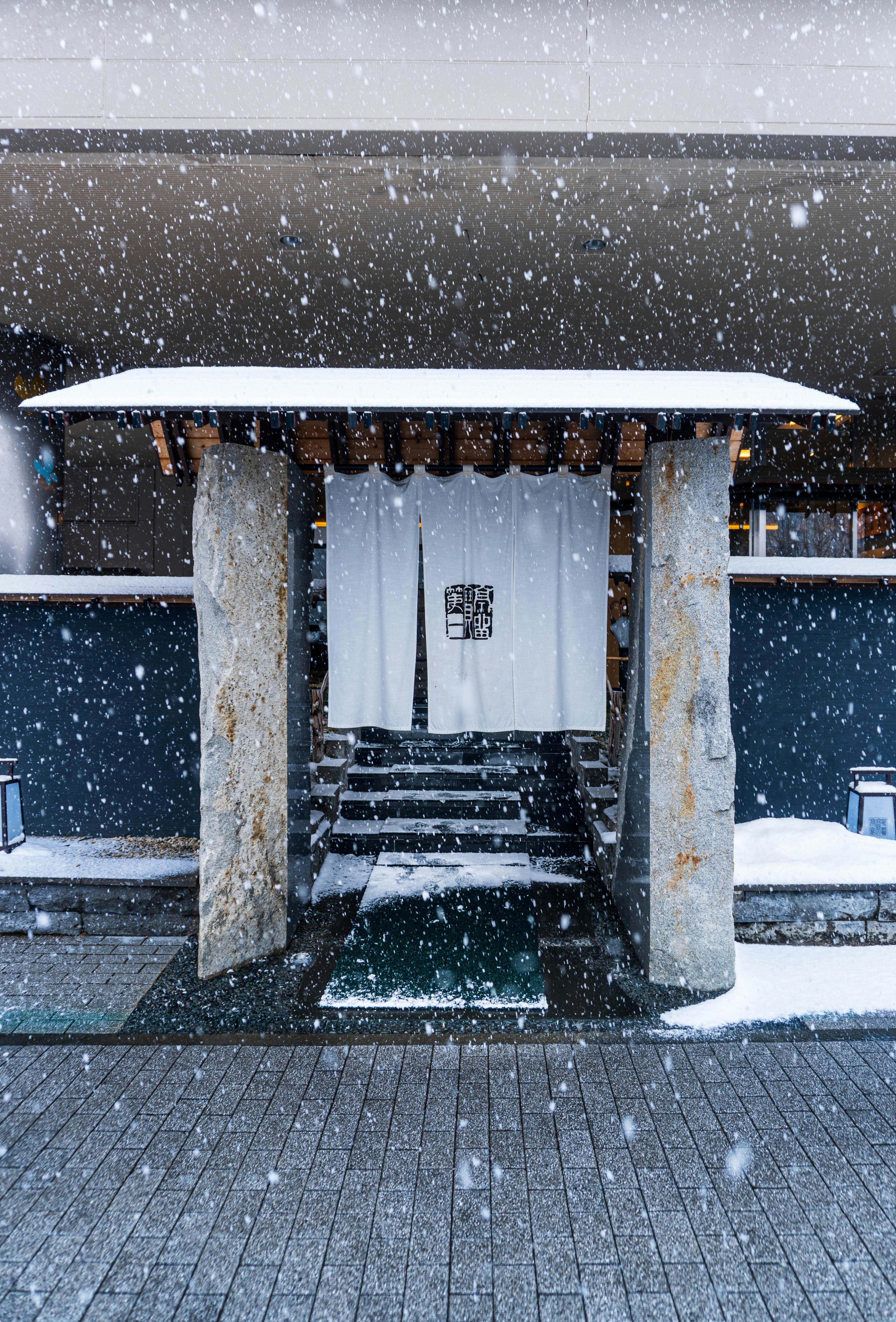 Traditional entrance adorned with a fabric curtain, framed by stone pillars, amidst a gentle snowfall.