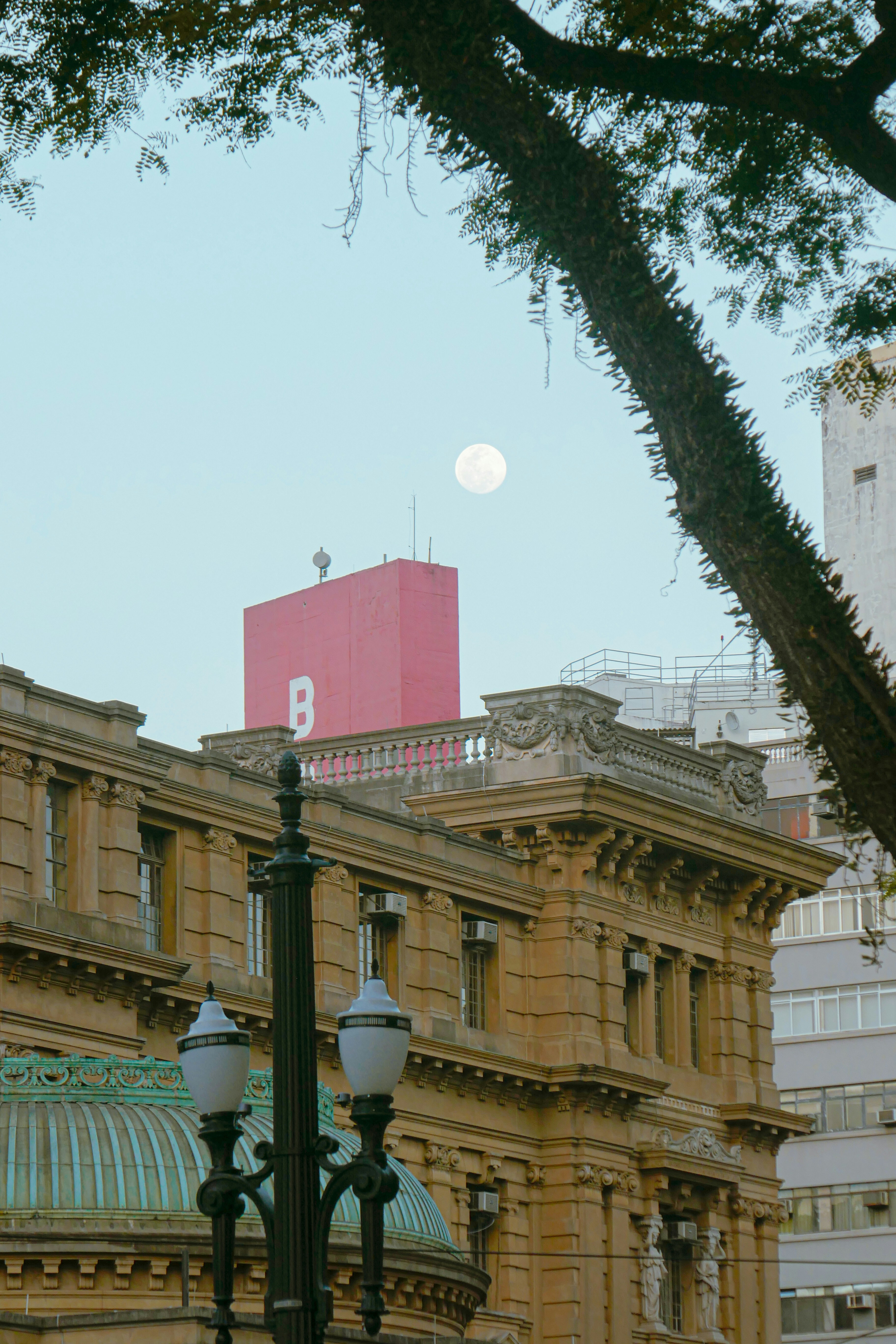 A full moon rises above a historic building, framed by lush foliage and modern structures in the background.