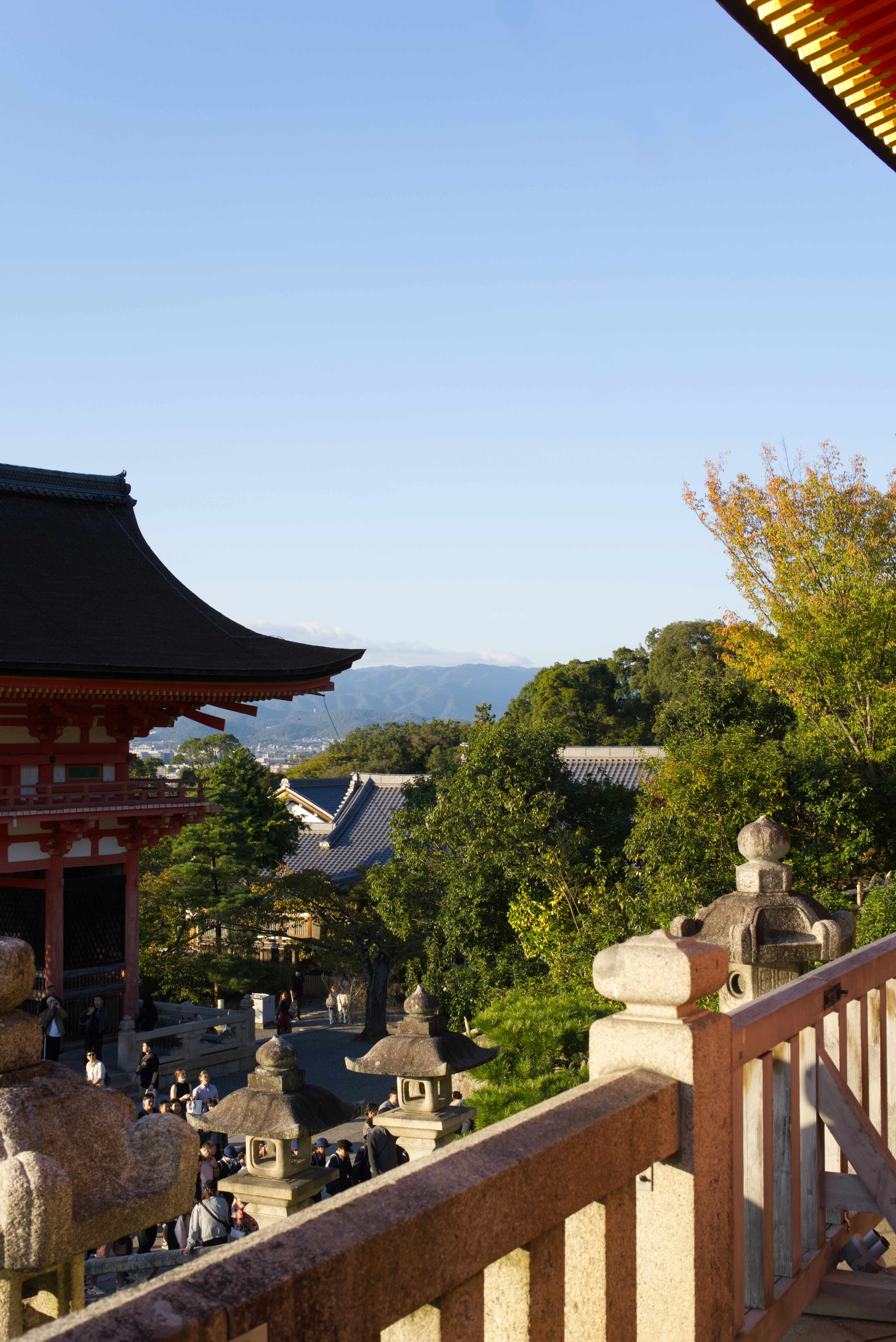 A tranquil view from a temple balcony overlooking lush greenery and traditional architecture, with distant mountains under a clear blue sky.