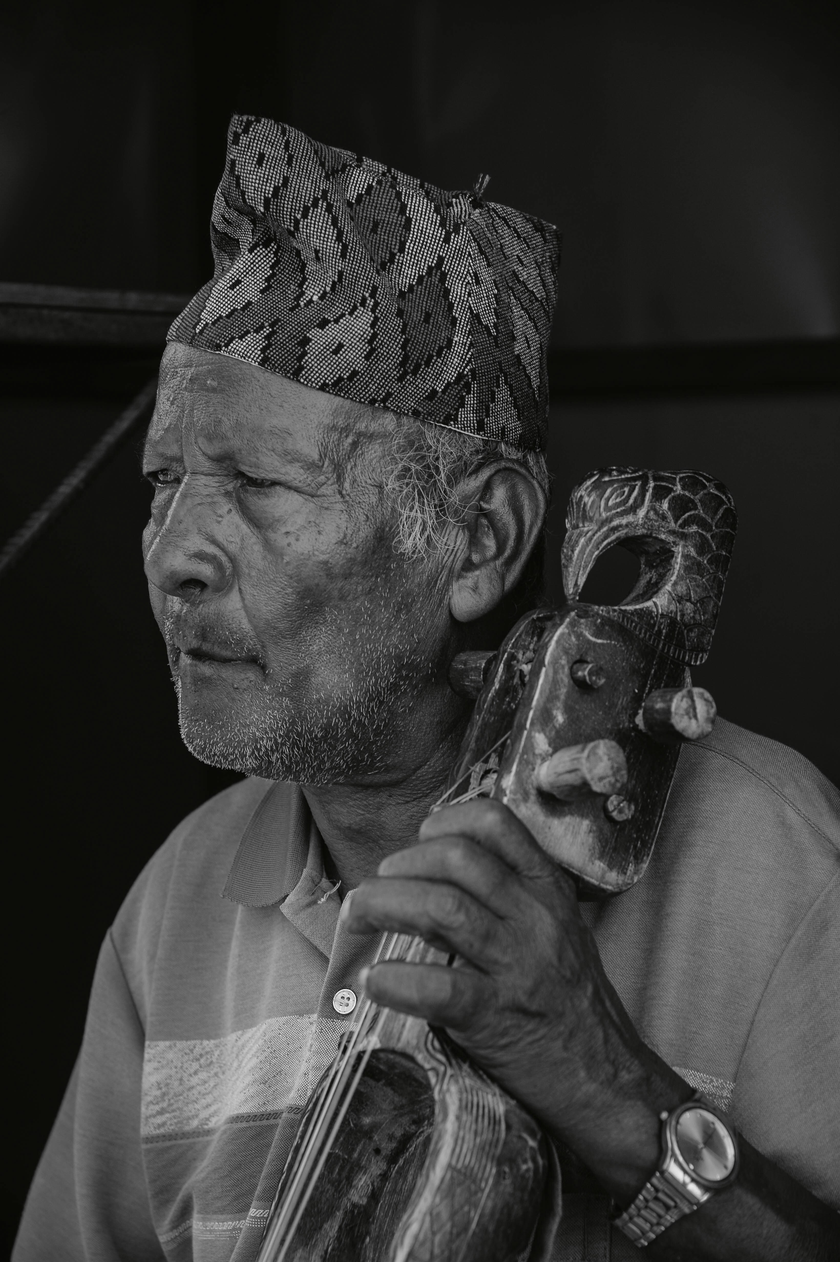 An elderly Nepali man plays a traditional sarangi with a warm, soulful expression. Captured in black and white, this portrait reflects the beauty of Nepali heritage, folk music, and the timeless connection between culture and emotion. His weathered hands and gentle smile tell stories of resilience, art, and life in the hills of Nepal. | Elderly man wearing a cap with a traditional instrument.