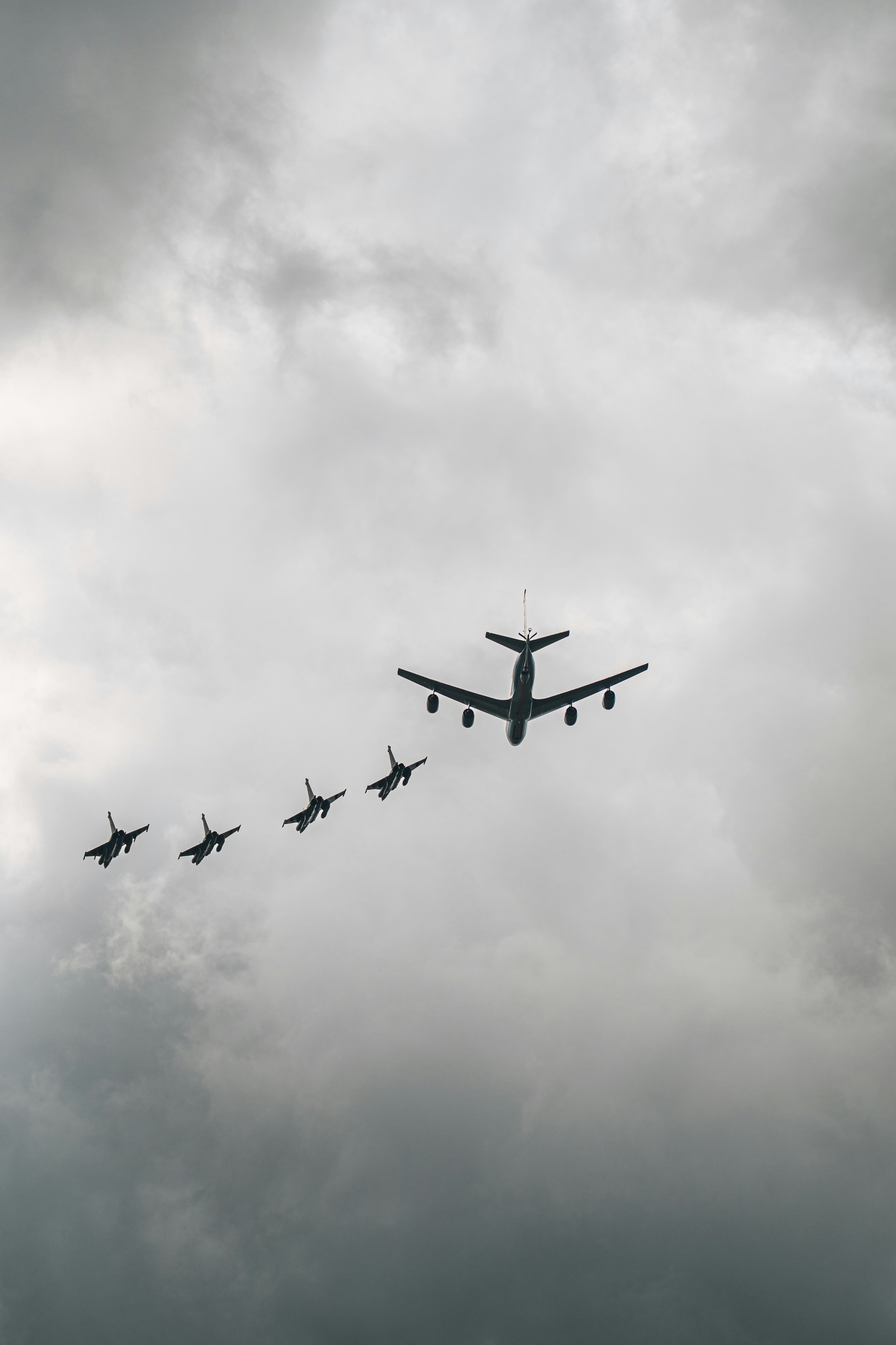 Formation of five fighter jets trailing behind a larger aircraft amidst a dramatic, cloudy sky.