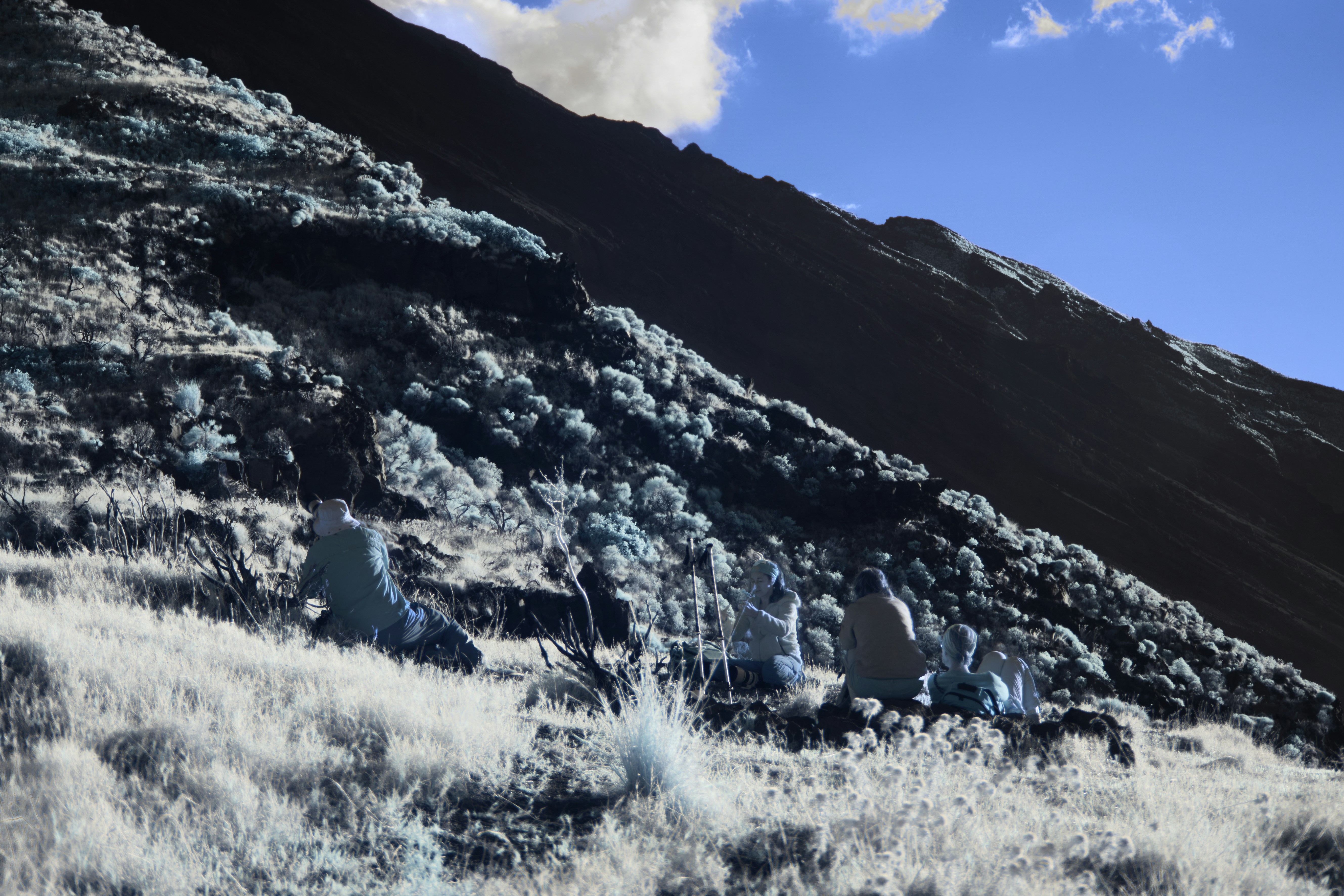 A group of individuals seated on a hillside, surrounded by lush vegetation and a dramatic mountain backdrop, captured in infrared tones.