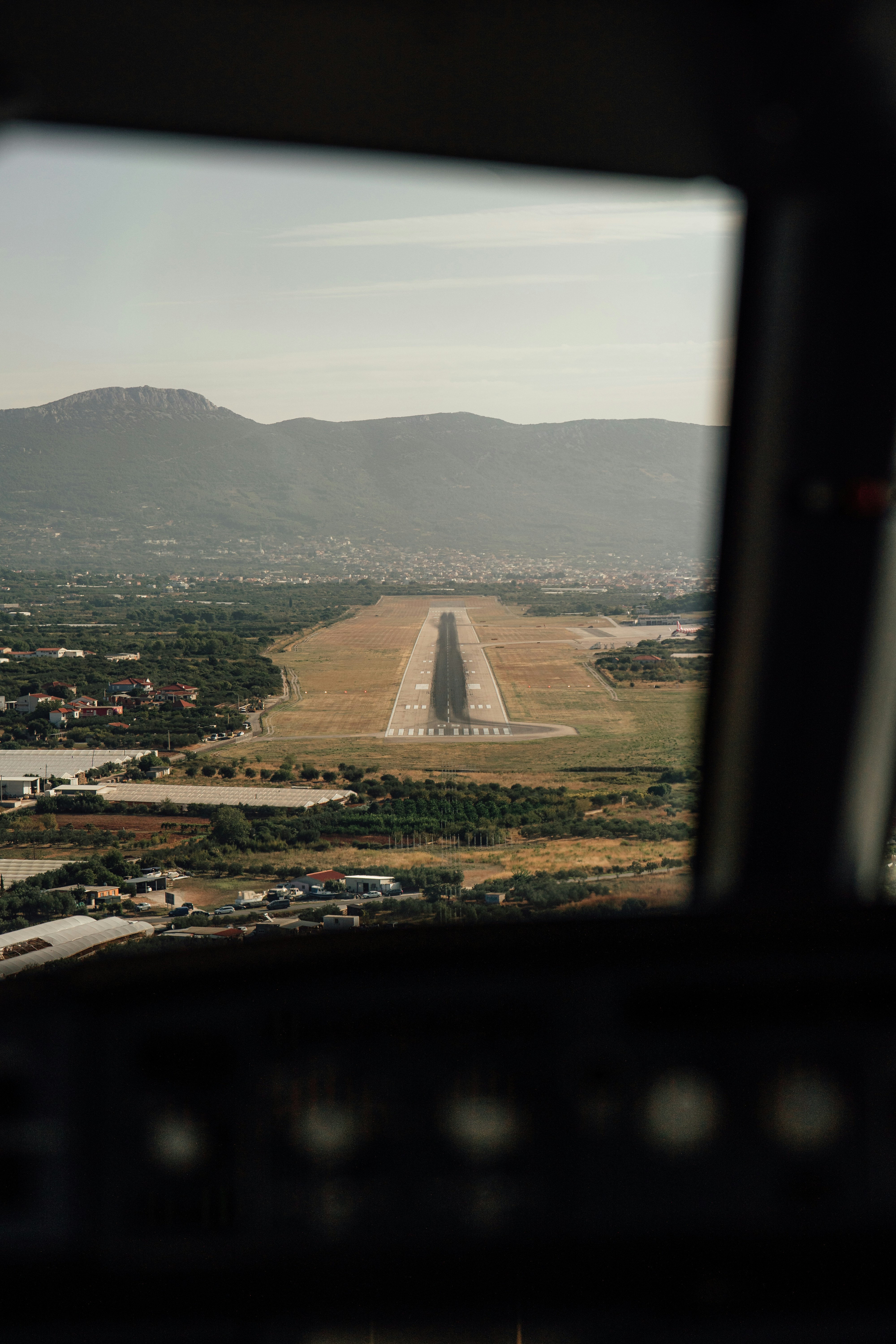 Runway stretching into the distance, framed by mountains and urban landscape, viewed from an aircraft cockpit.