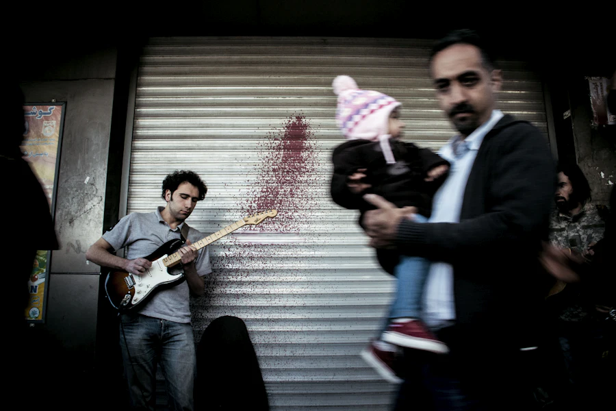 A young man plays an electric guitar against a metal shutter marked by red paint, while a blurred man carrying a child passes in the foreground. The scene feels like candid street life with motion and contrast.