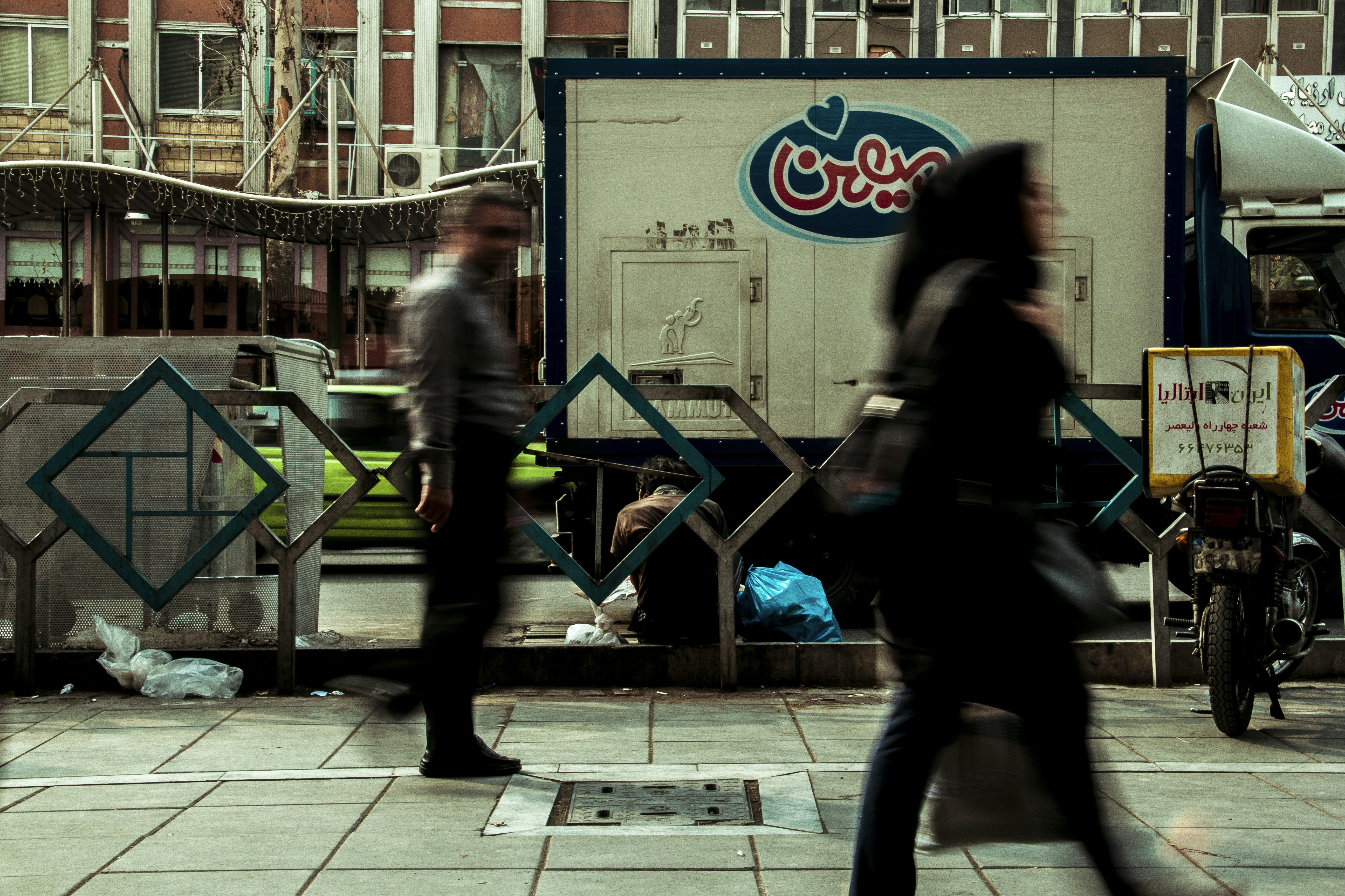 Blurred figures navigate a bustling street, with a colorful truck serving as a backdrop, capturing the essence of urban life and movement.