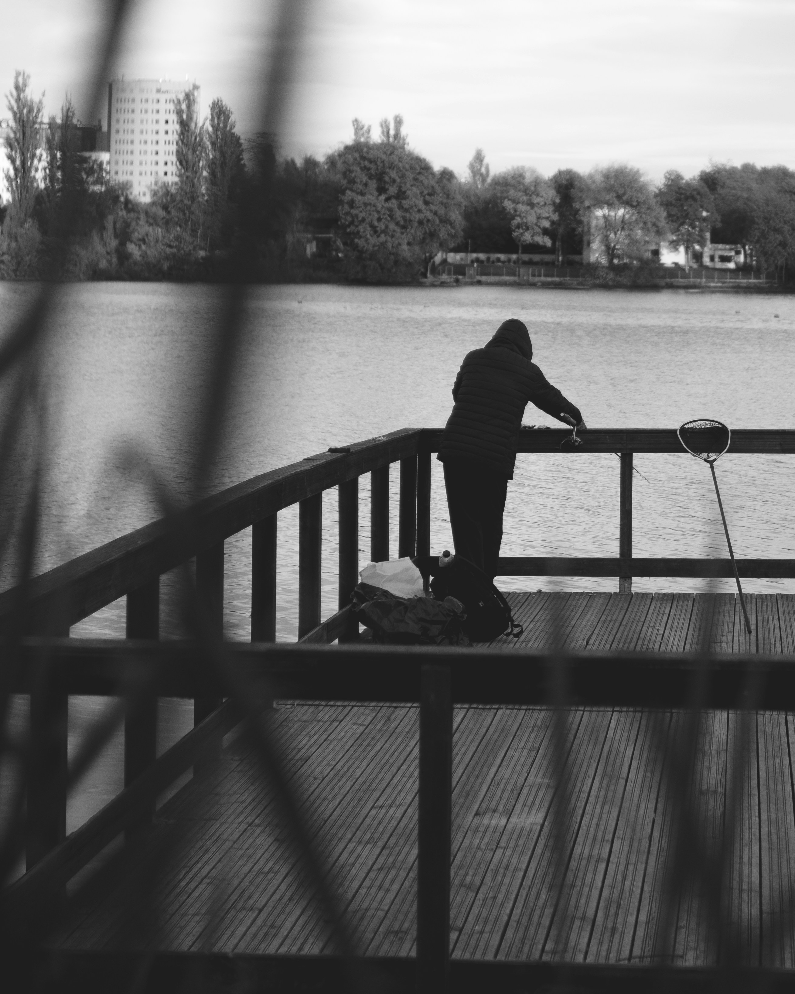 Person leaning over a wooden pier by a tranquil lake, surrounded by trees and urban skyline in the background.