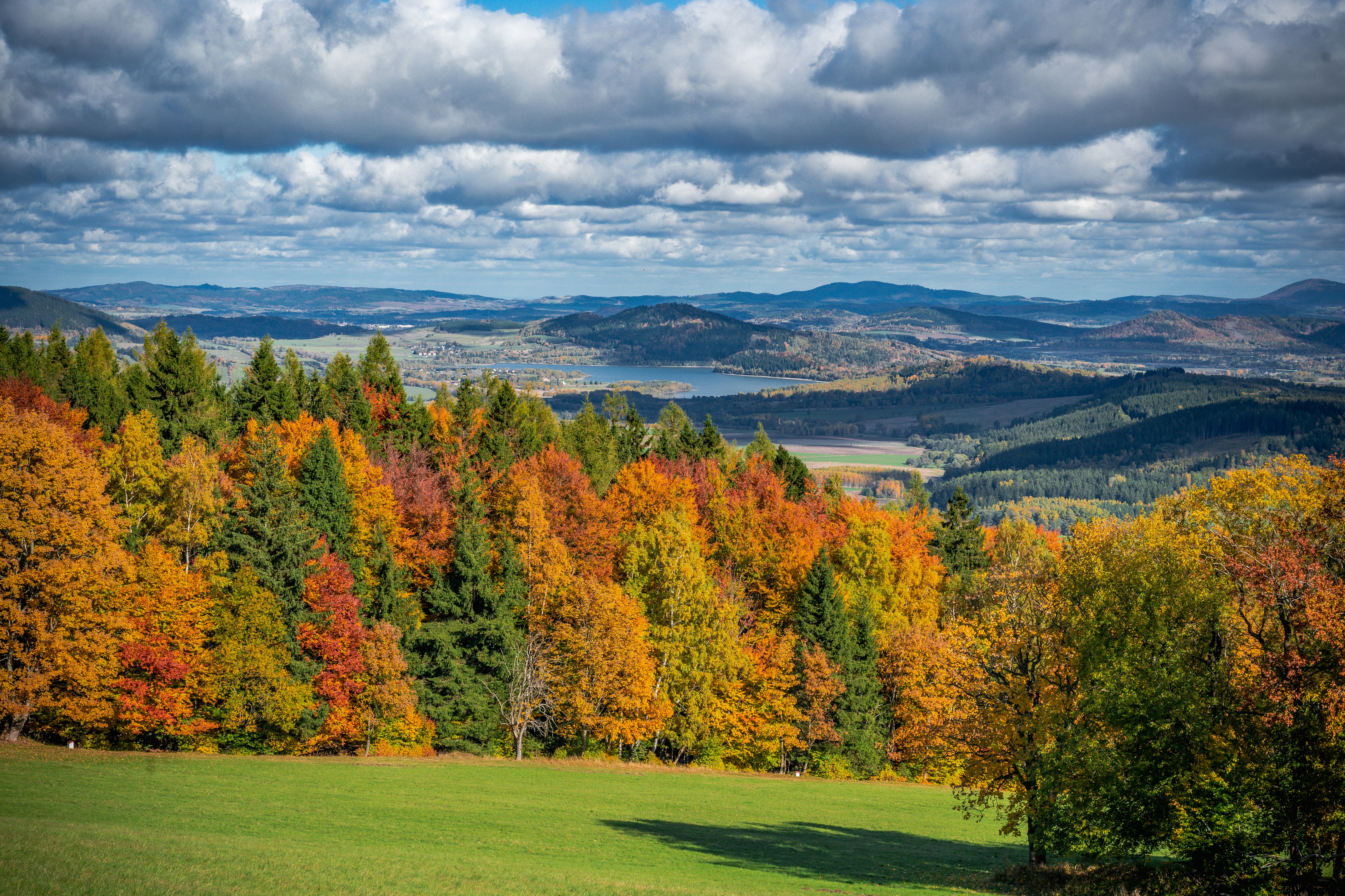 Vibrant autumn foliage blankets the hills, contrasting with the serene lake and distant mountains under a dynamic sky.