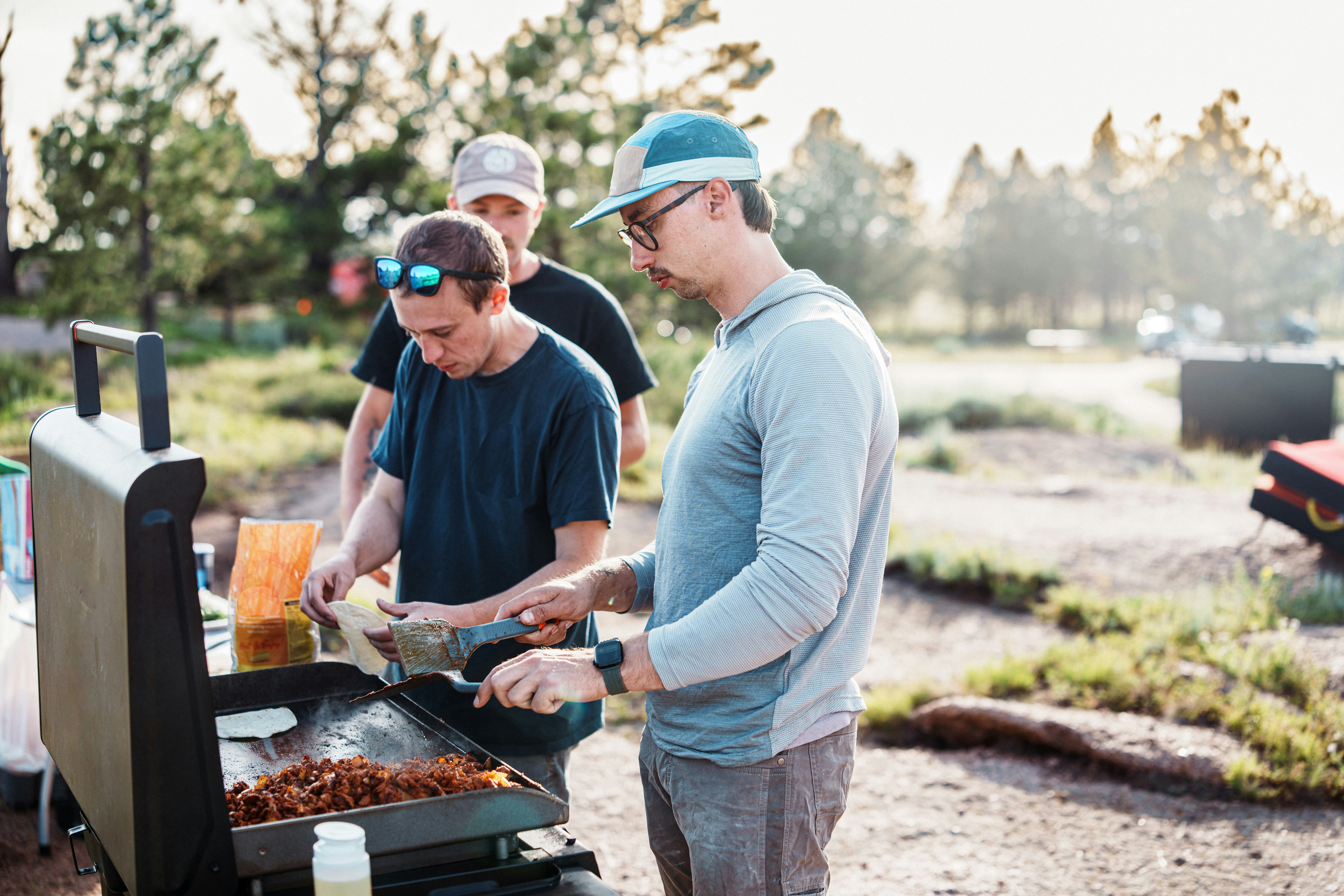 Three friends gather around a grill, preparing a meal in a sunlit outdoor setting. The scene captures the essence of friendship and outdoor cooking.
