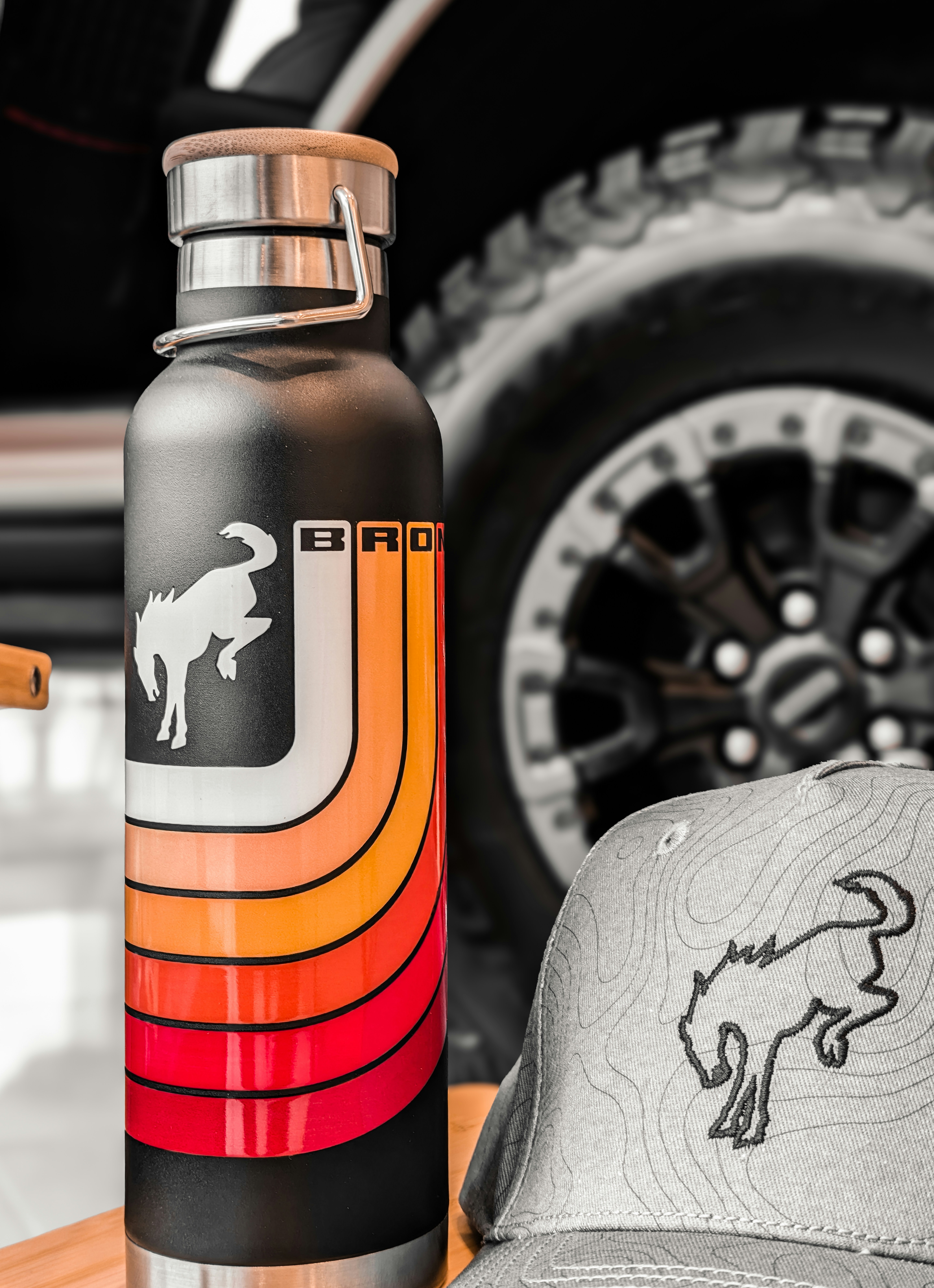 Stylish water bottle featuring a vintage Bronco logo, accompanied by a matching hat, set against a blurred backdrop of rugged tires.