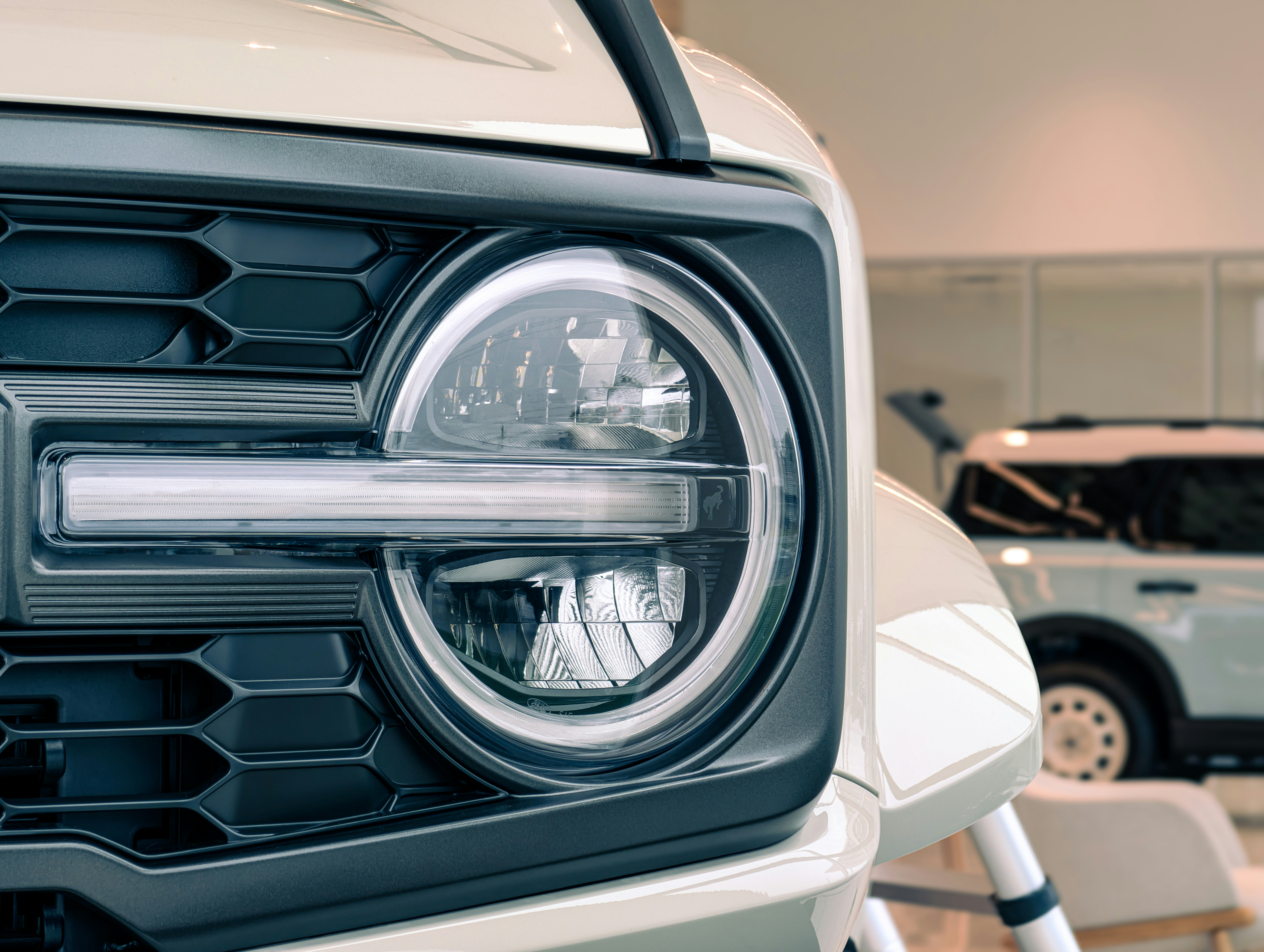 This bright, ultra-clean close-up captures the signature, retro-modern gaze of the Ford Bronco headlight, a magnificent homage to its rugged heritage. The powerful LED ring and bold horizontal Daytime Running Light (DRL) bar flawlessly bisect the classic round lens, creating a striking visual tension against the aggressive, honeycomb grille texture. The light-toned body panel in the high-key environment adds a luminous, sophisticated polish to this piece of iconic automotive design. | Close-up of a modern suv's headlight and grille.