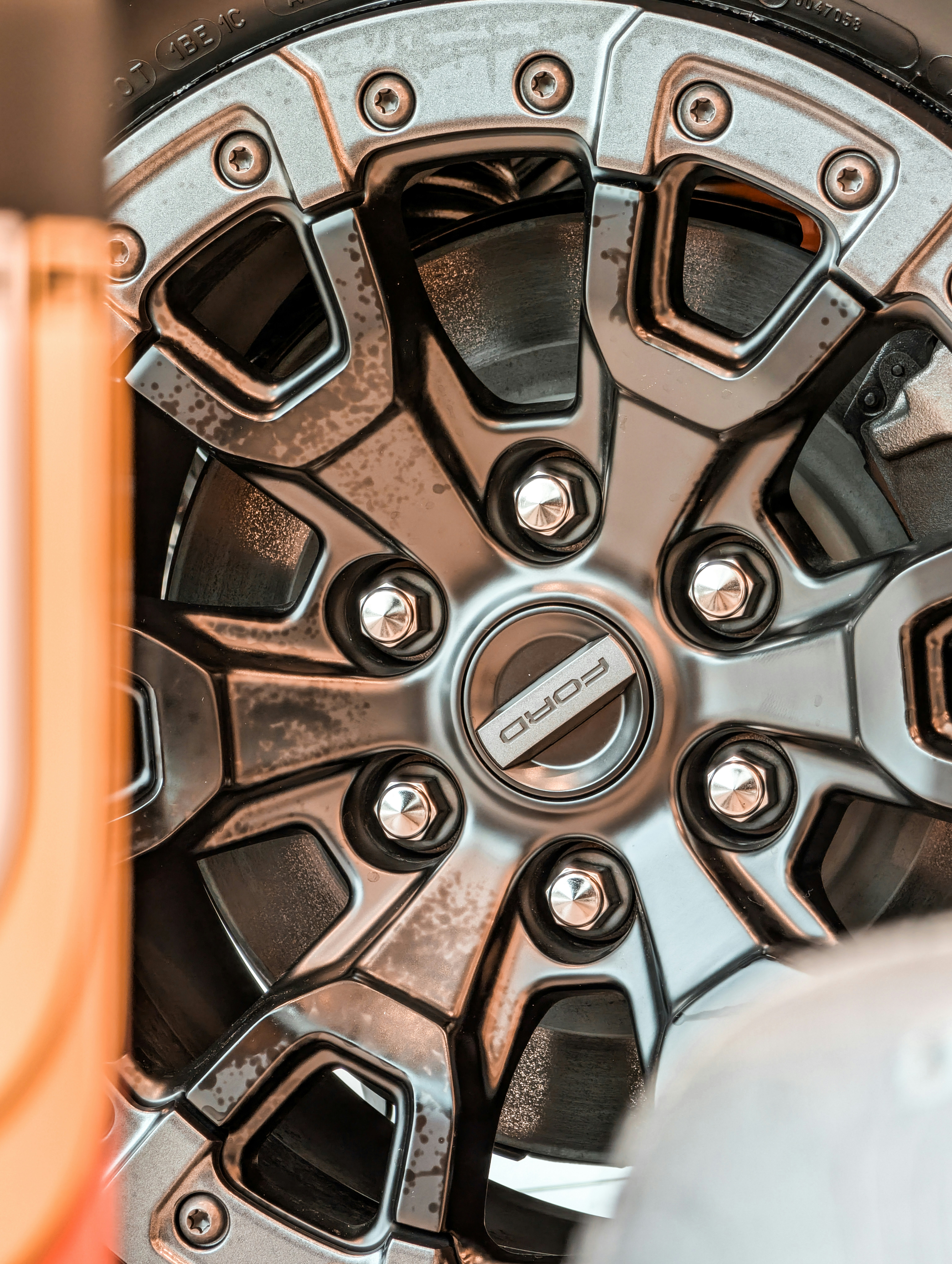 This striking, portrait-oriented close-up celebrates the sheer engineered toughness of the Ford Bronco wheel, a visual masterpiece of rugged design. The complex, multi-spoke alloy, with its technical beadlock ring secured by gleaming lug nuts, captures a beautiful, moody patina that contrasts sharply with the reflective gloss. A soft, warm orange glow, likely a reflection, subtly bathes the wheel, highlighting its powerful, sophisticated aesthetic ready for any extreme challenge. Boulevard Ford in Millsboro