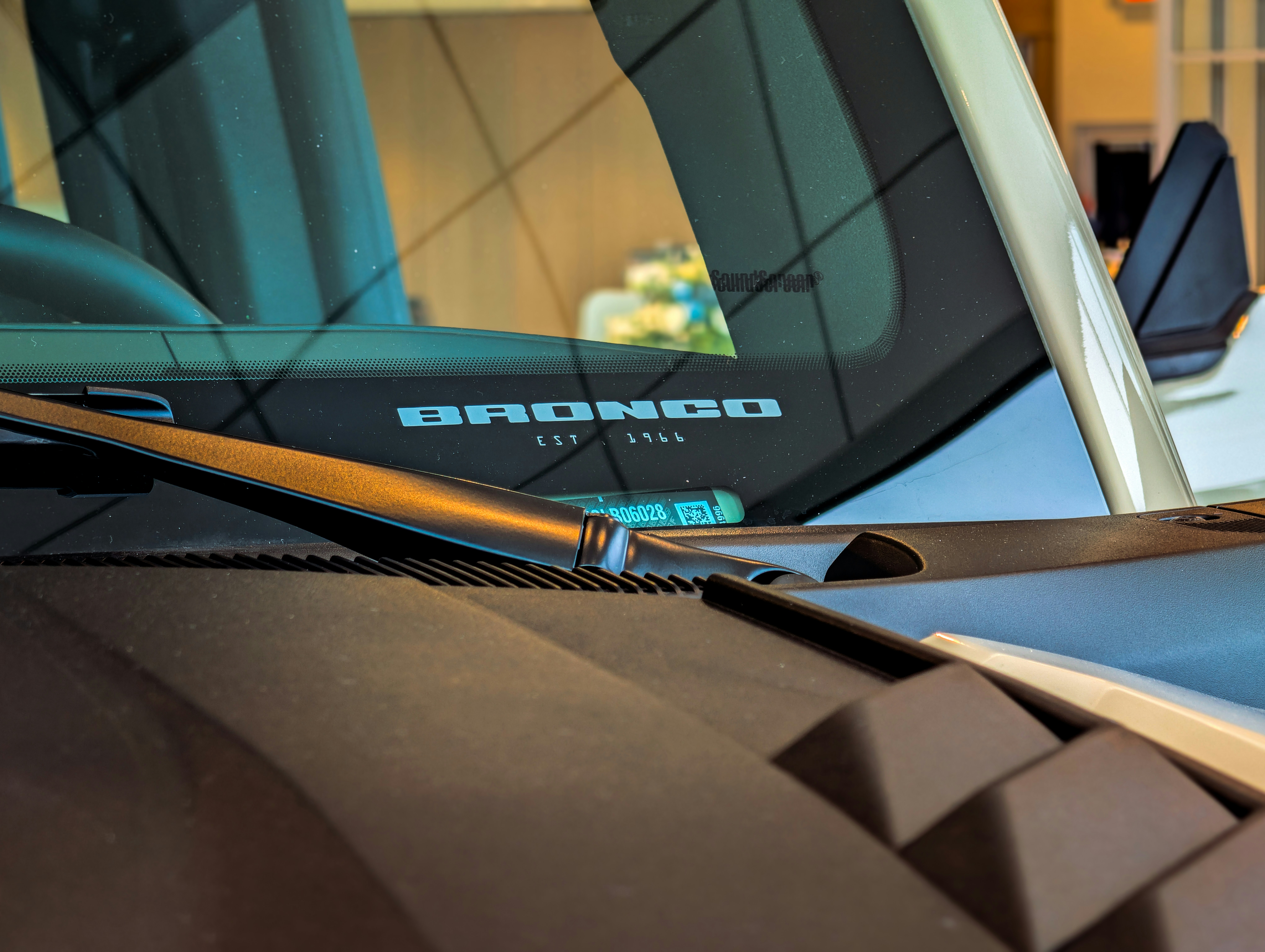 This sharply focused composition showcases a proud, yet subtle nod to history, with the "BRONCO EST 1966" lettering flawlessly etched into the upper windshield glass. The elegant white text stands as a powerful statement of heritage against the deep, dark reflection of the angled glass and the textured black hood. The striking contrast between the smooth glass and the rugged utility of the surrounding materials beautifully embodies the fusion of legacy and cutting-edge design. | Bronco logo on a car windshield