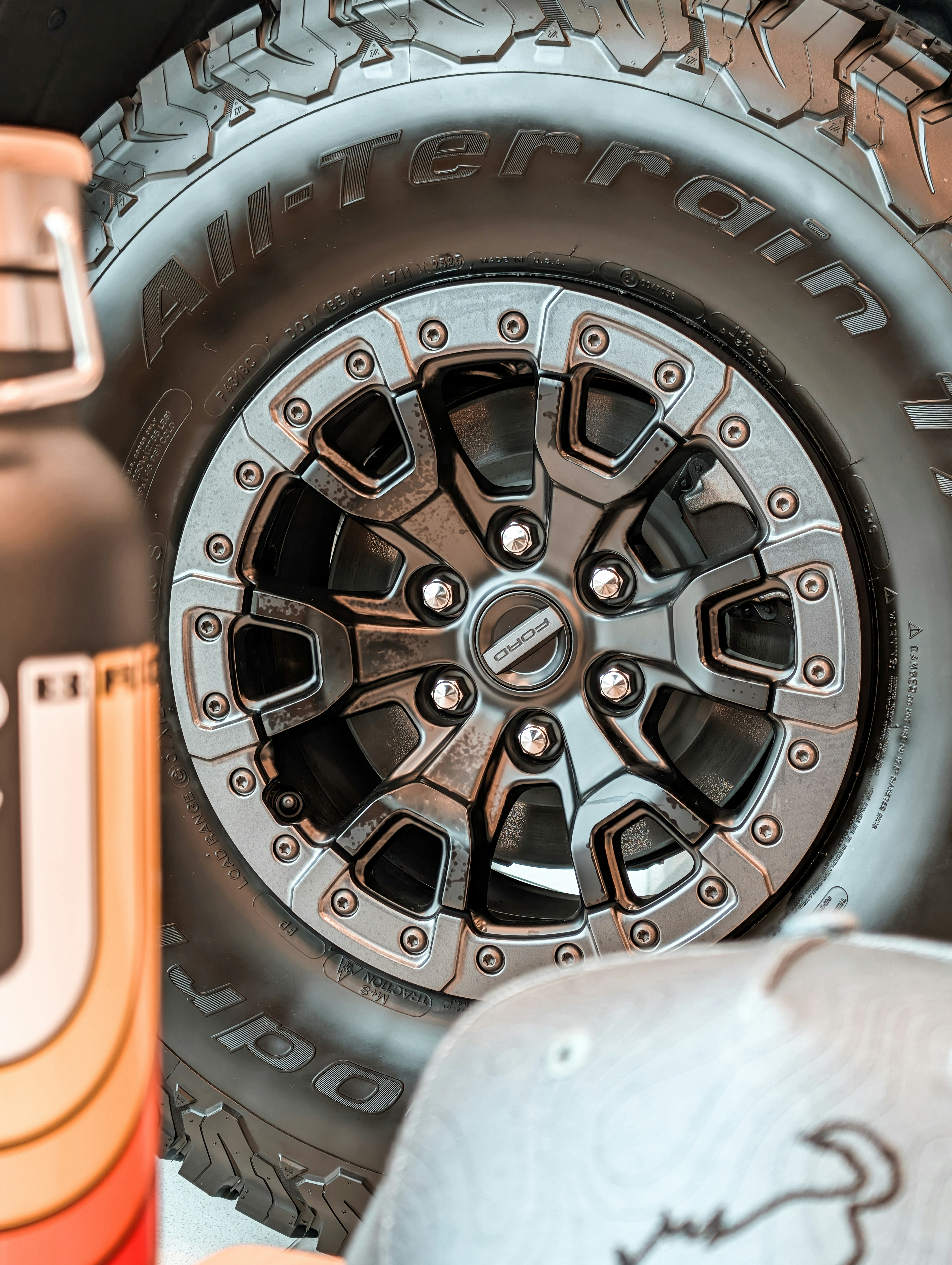 Close-up of a rugged all-terrain tire and alloy wheel, showcasing intricate design and durability. A water bottle and cap are partially visible in the foreground.