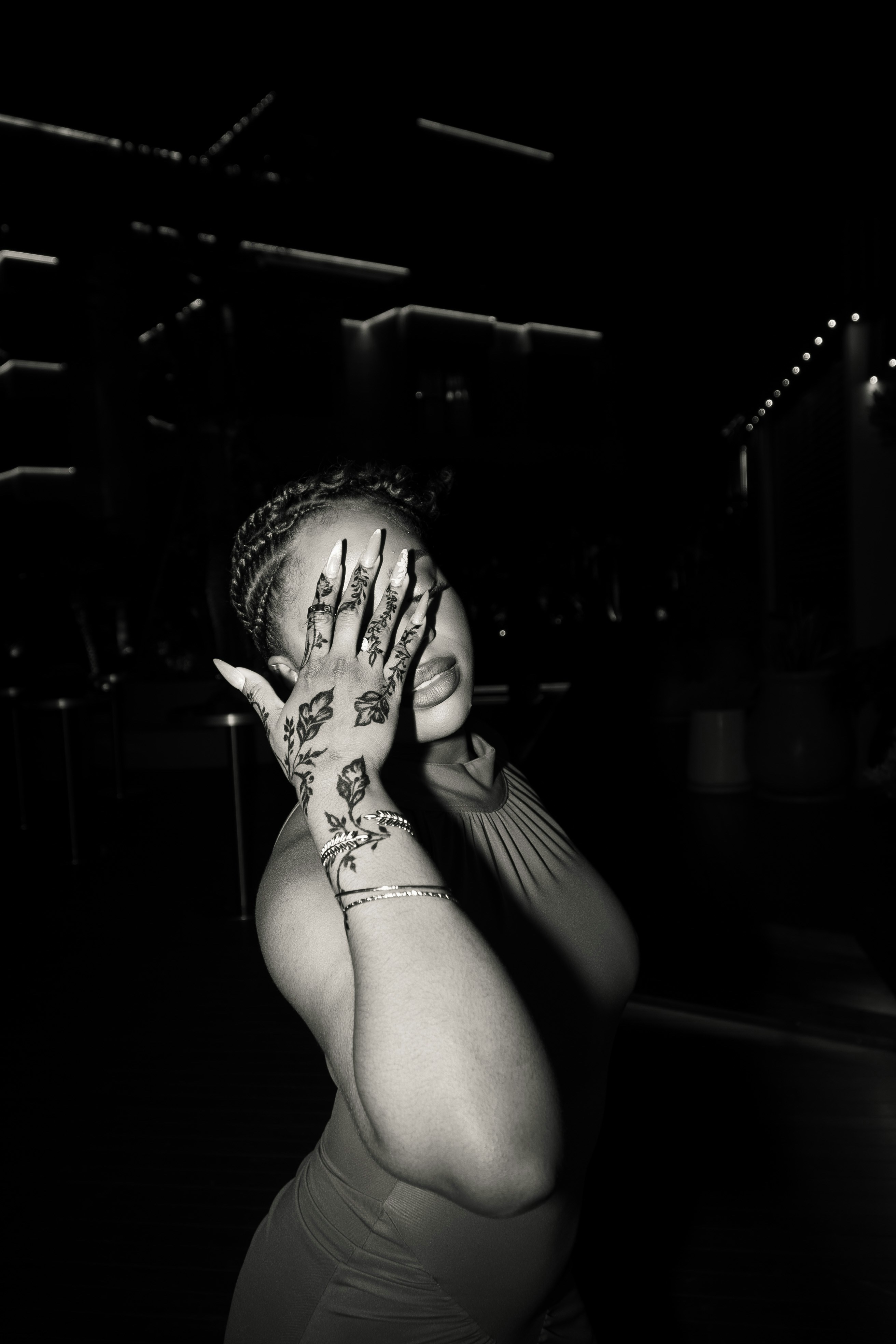 A woman elegantly obscures her face with intricately tattooed hands, set against a dimly lit backdrop. The contrasting light enhances the depth of the scene.
