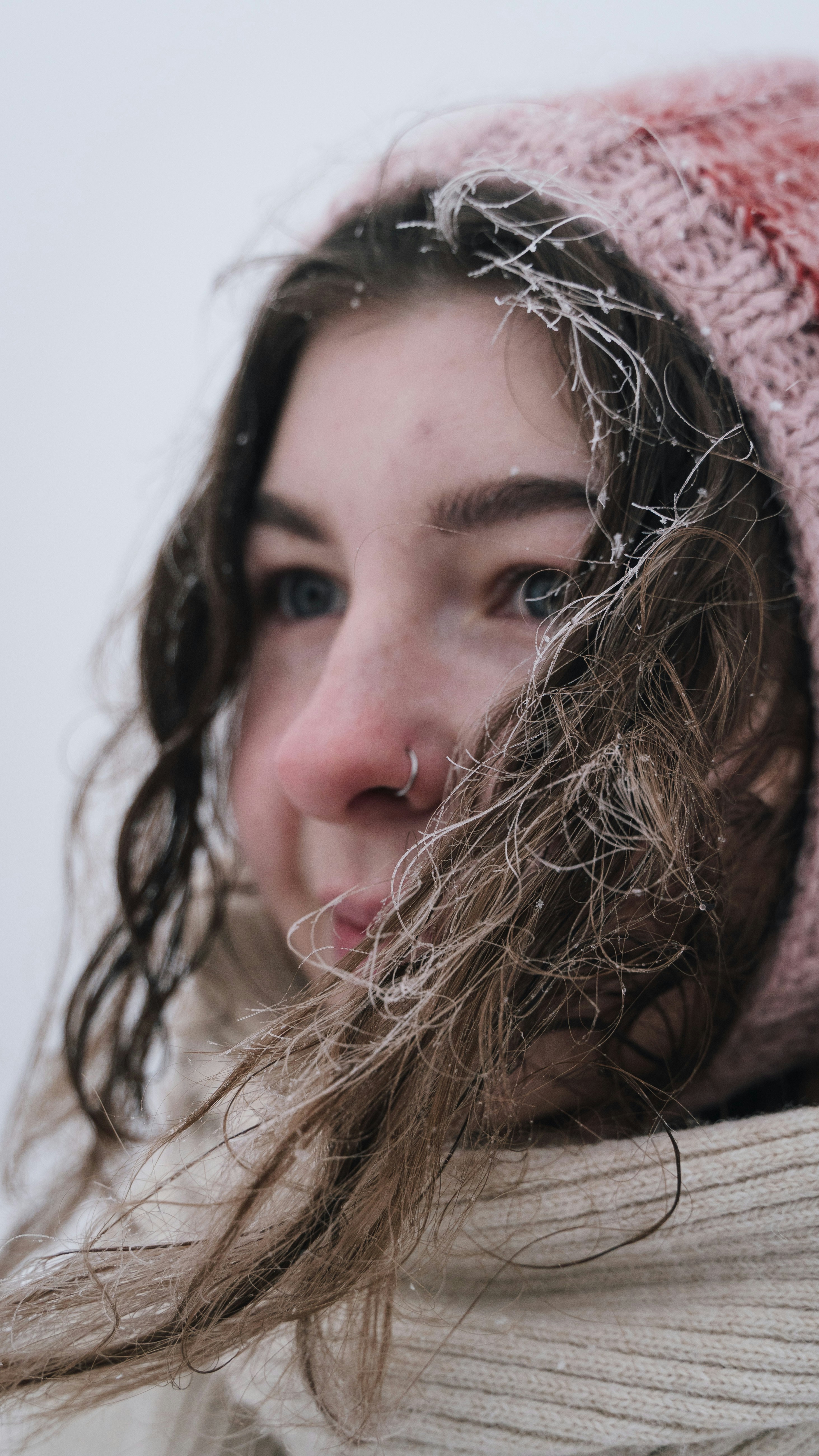 Close-up of a young woman with wind-tousled hair and frost-kissed strands, wearing a cozy pink beanie and scarf against a snowy backdrop.