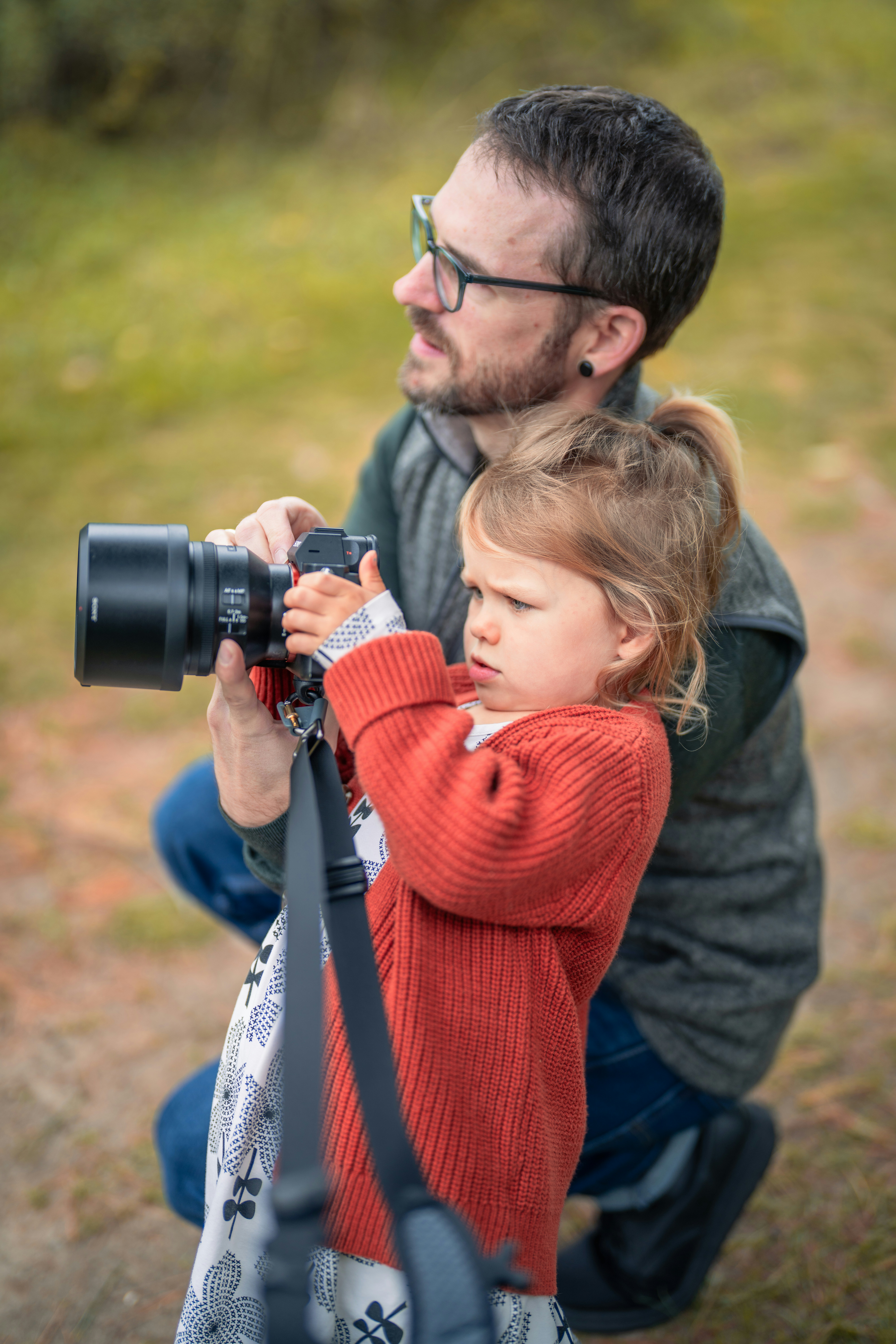 A young child gripping a camera while observing a scene, guided by an adult. The focus is on the child's expression and engagement with photography.