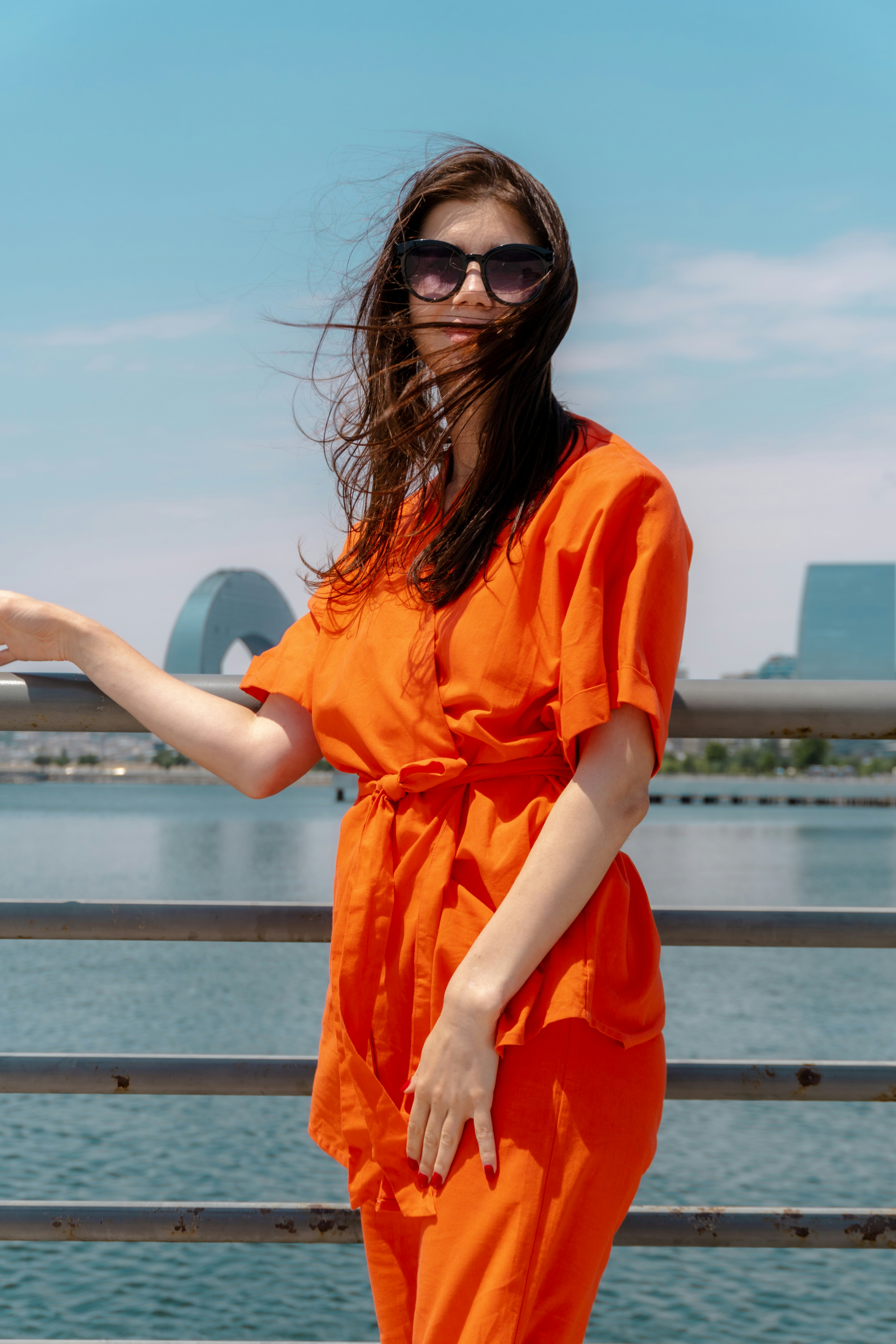 Woman in a vibrant orange outfit stands by a railing with a scenic waterfront backdrop. Her hair flows in the breeze, adding a dynamic element to the composition.