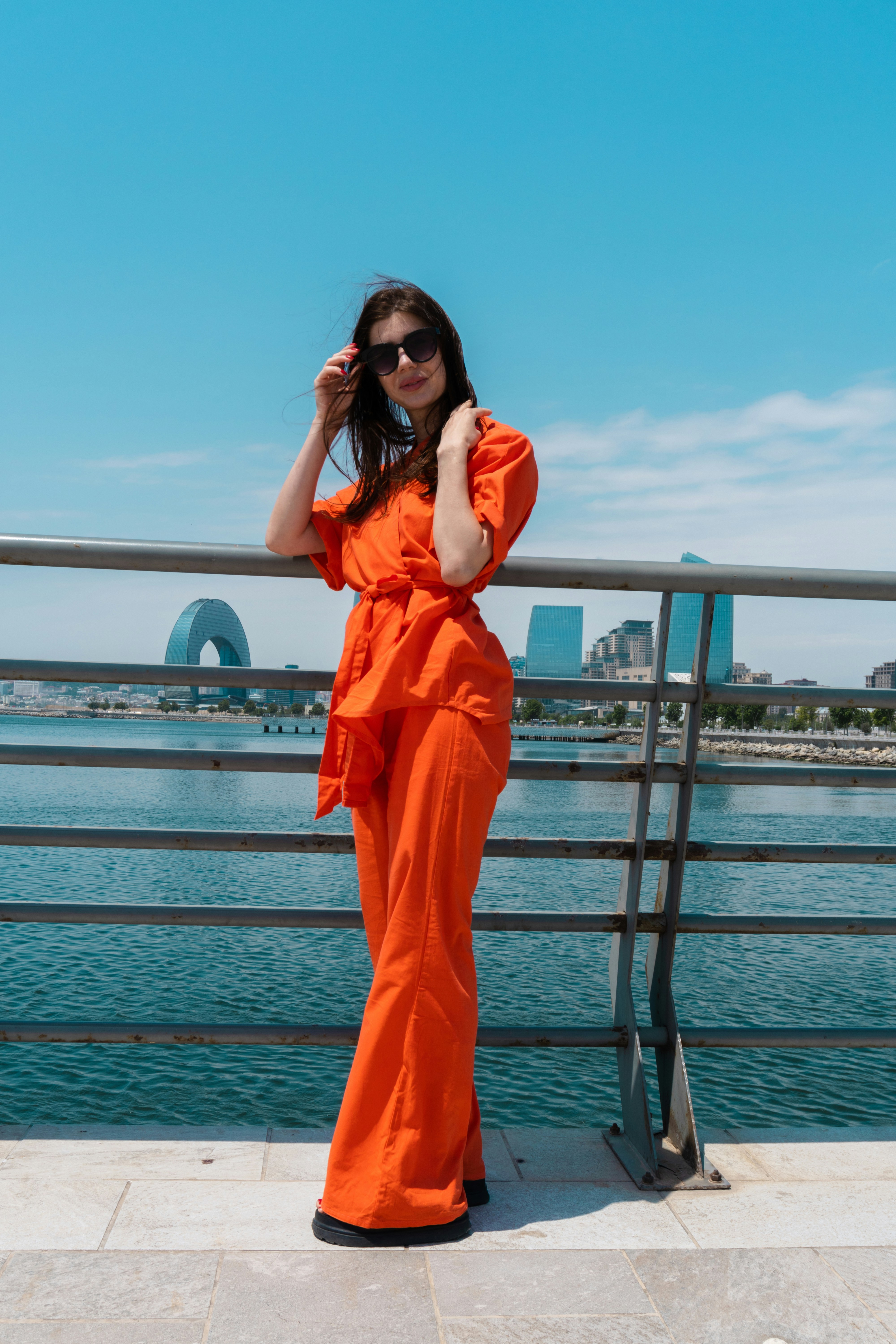 Woman in an orange outfit striking a pose against a scenic waterfront backdrop with modern architecture in view.