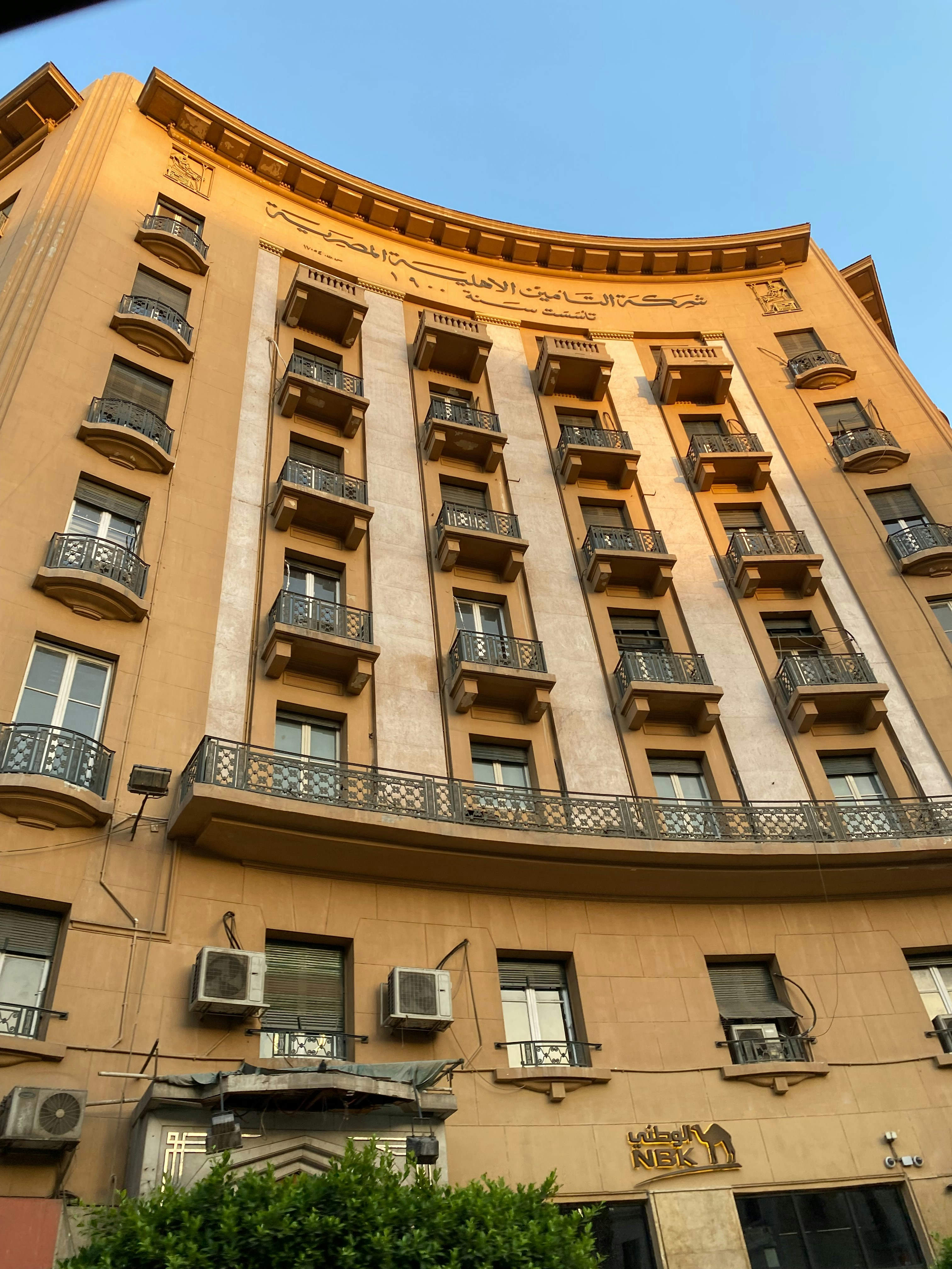 A historic building with a gracefully curved facade, featuring multiple balconies and intricate architectural details. The warm tones of the structure contrast with the clear blue sky.